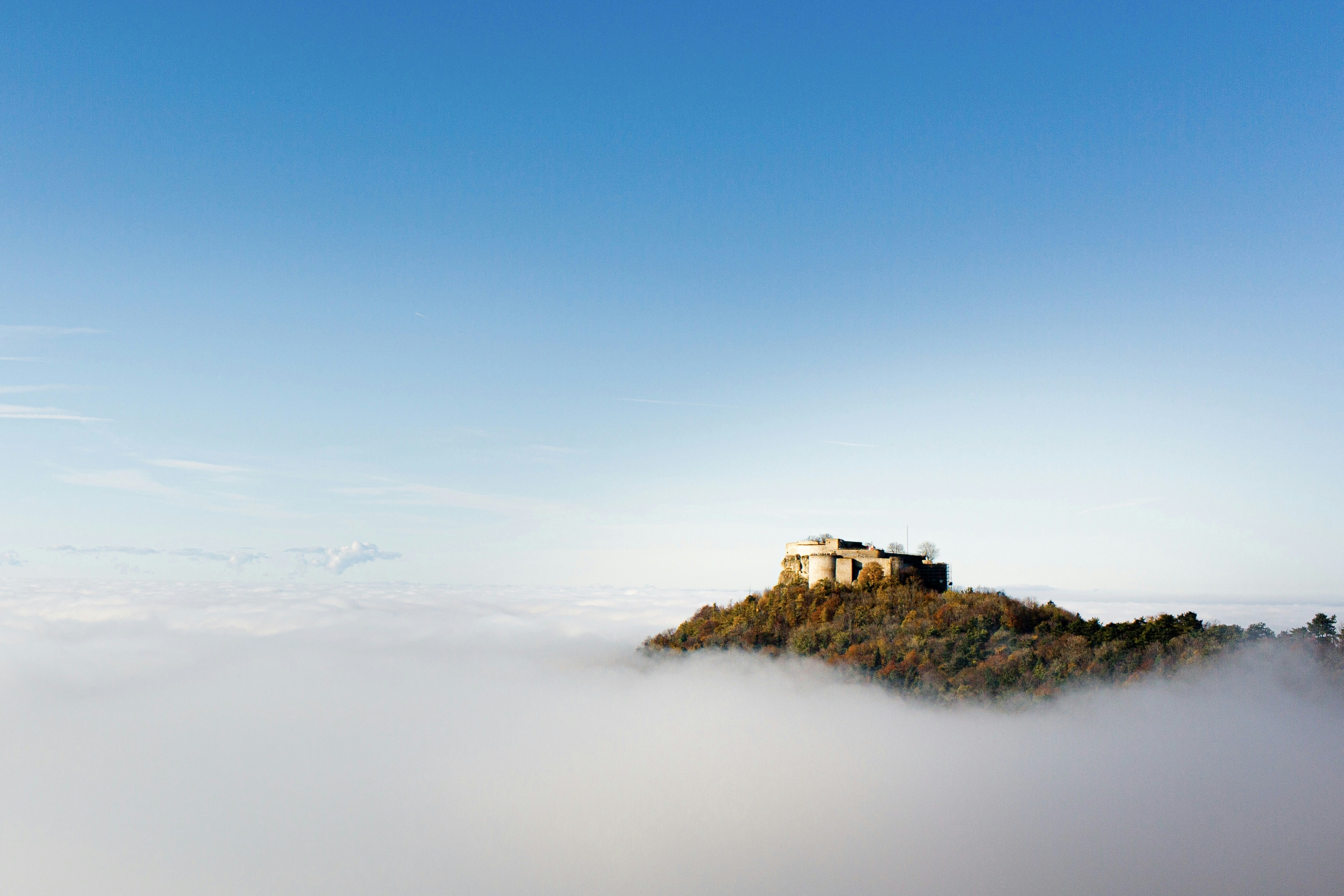 Ancient fortress perched on a hill, surrounded by a sea of fog under a clear blue sky.