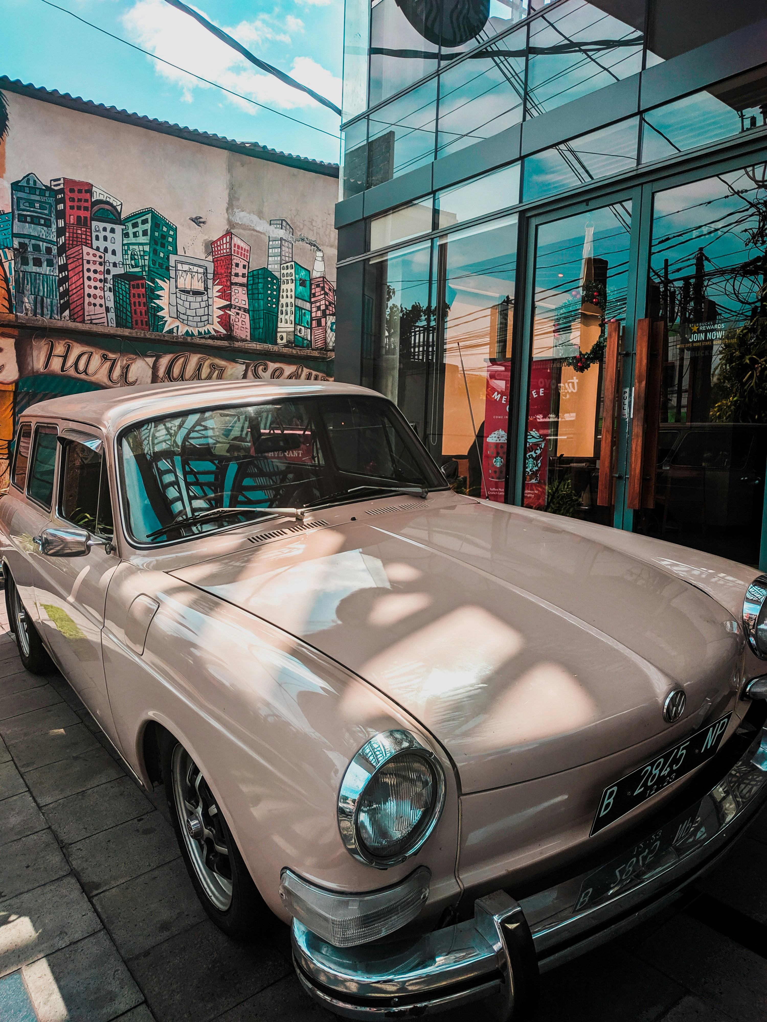 Classic pink car parked in front of a vibrant mural depicting colorful buildings and city life.