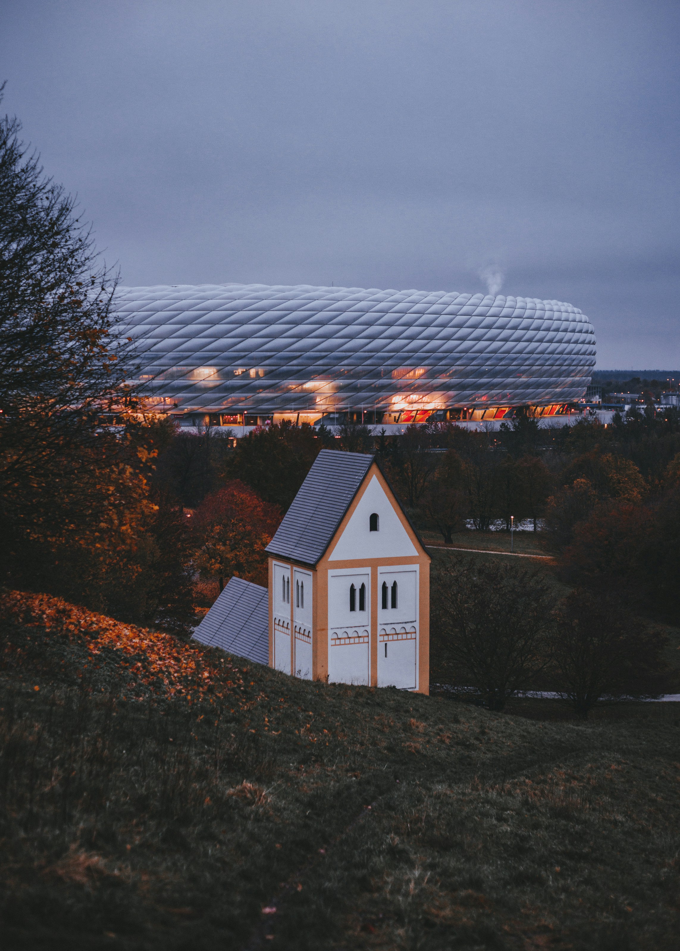 Dusk photograph of a white hillside house with a slate roof, framed by autumn trees, with a glowing ribbed stadium behind.