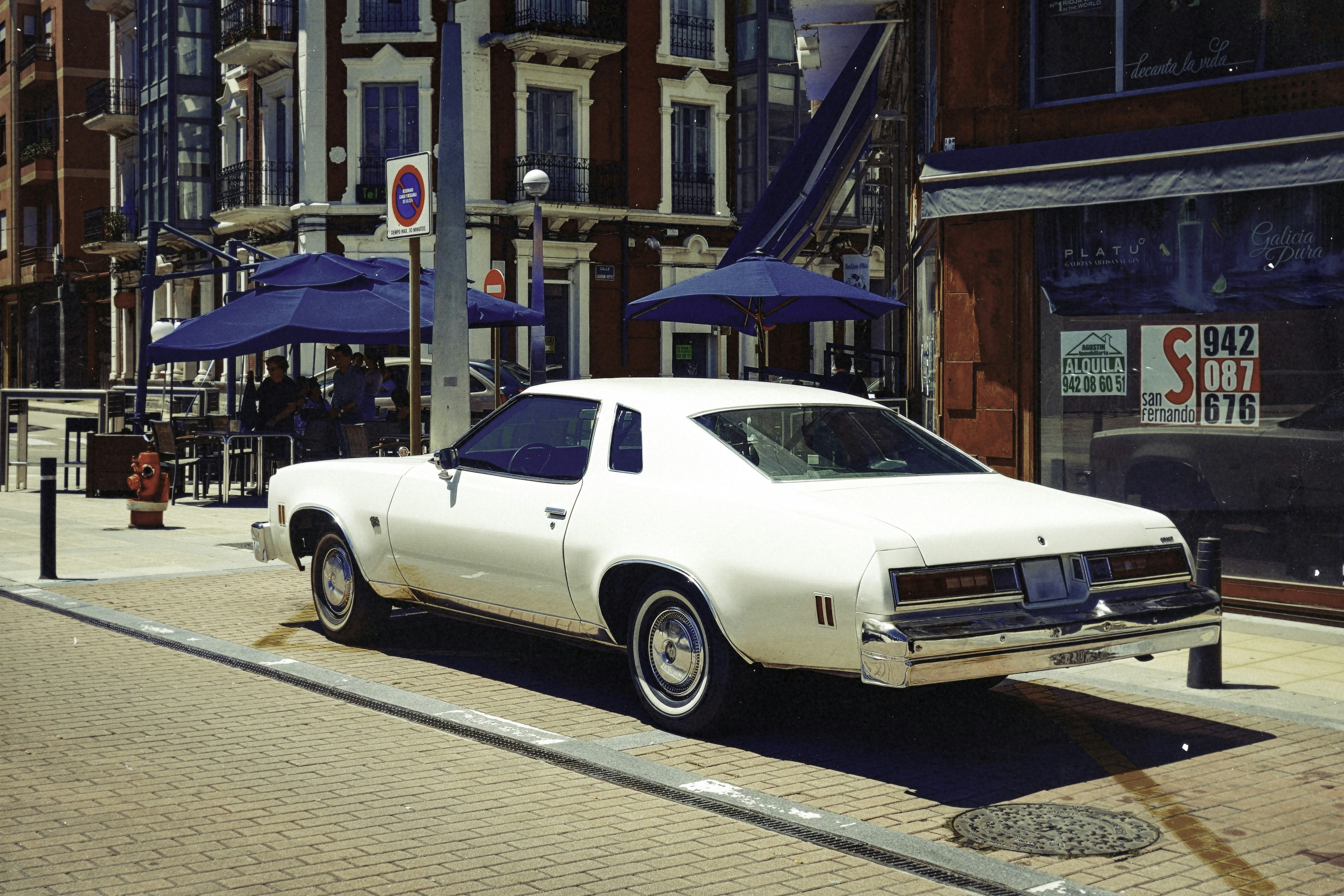 Vintage white coupe parked near vibrant urban cafes under clear skies.