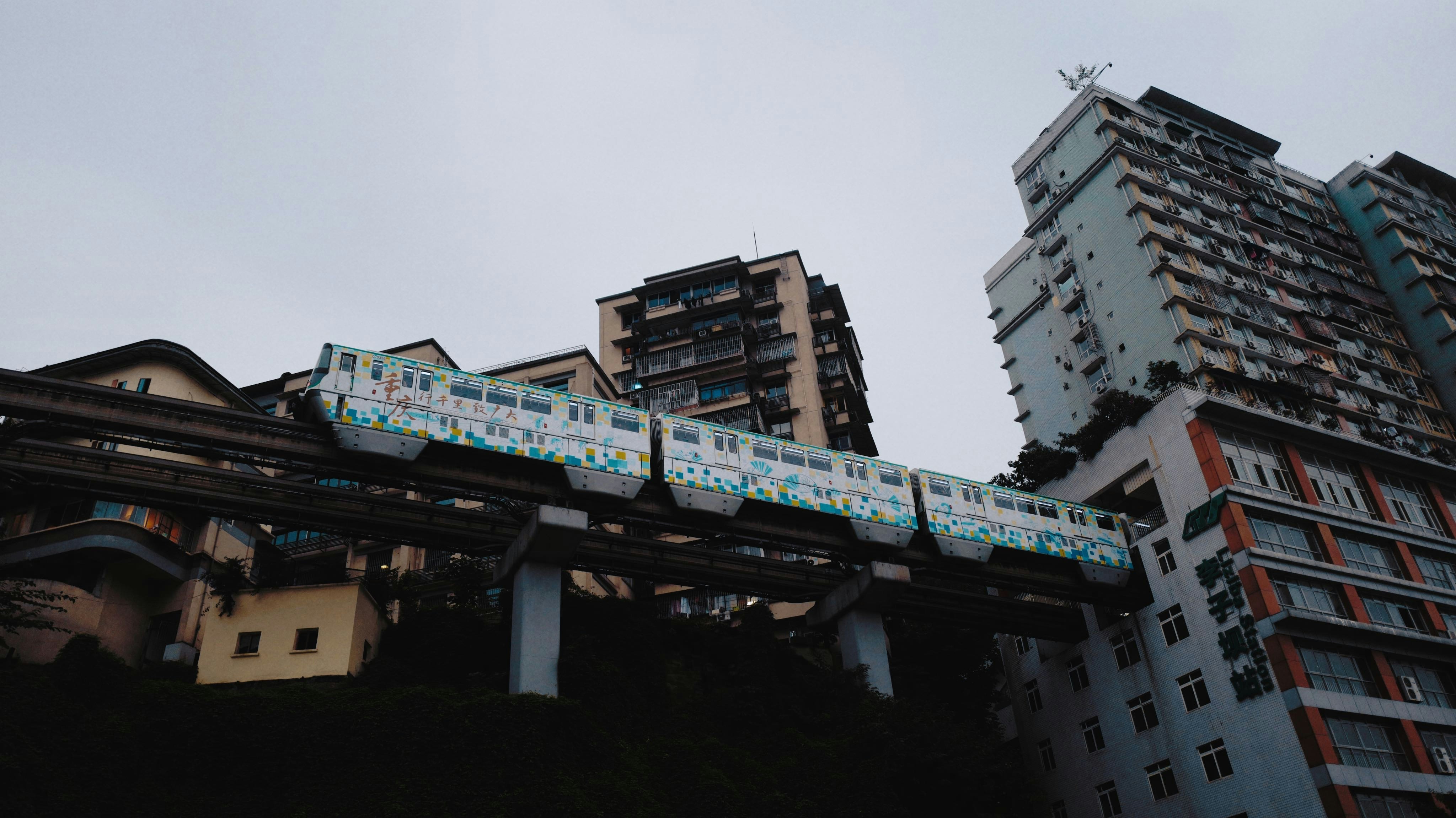 A monorail train glides above a hillside neighborhood, juxtaposed against towering buildings under an overcast sky.