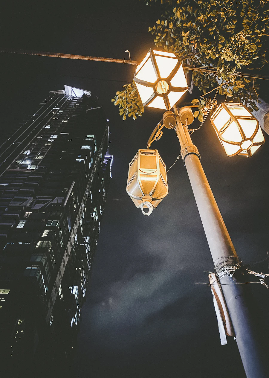 Warm geometric street lamps glow beside a tall dark skyscraper under a night sky. Tree leaves frame the upper right corner.