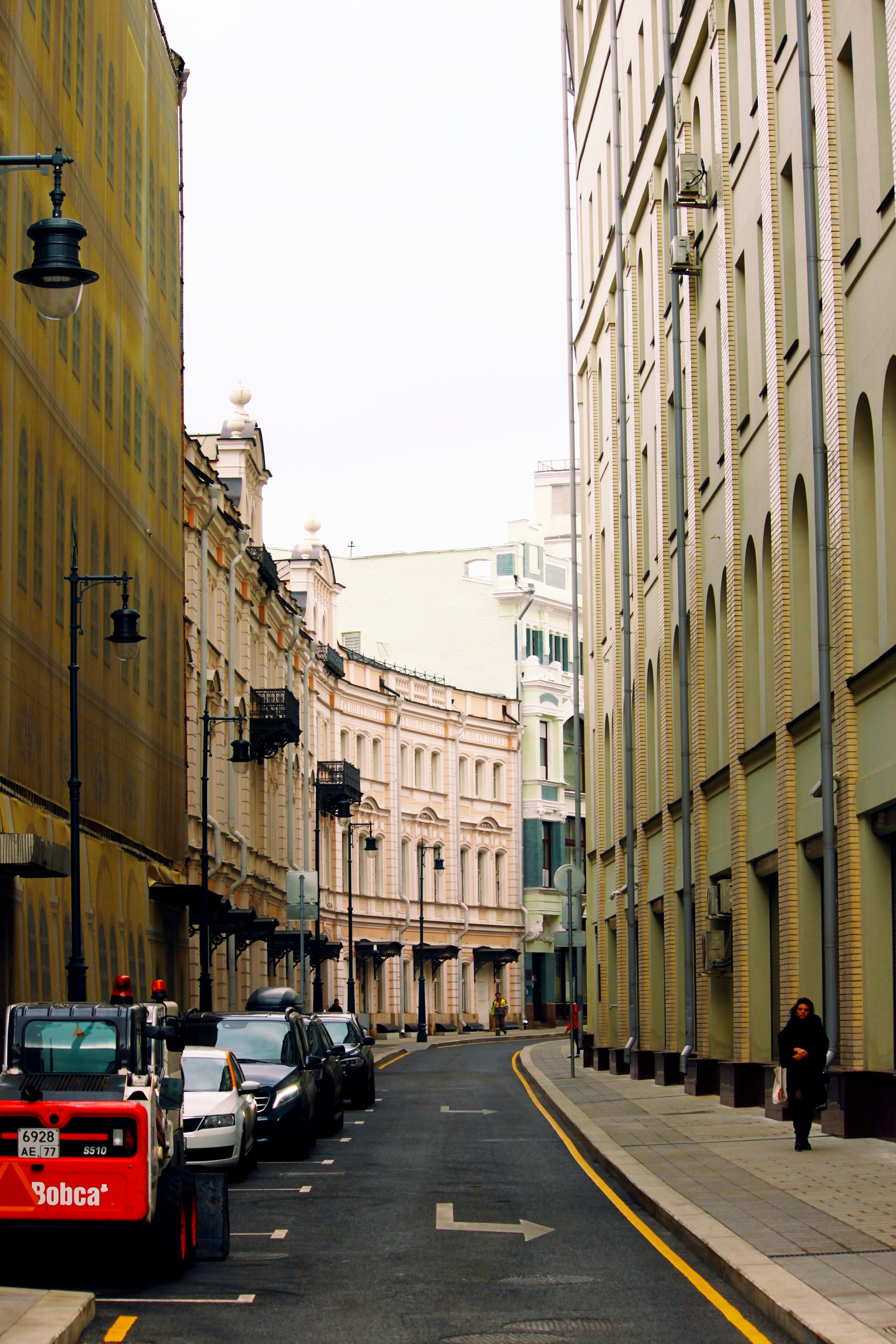 Narrow city street flanked by historical and modern buildings, showcasing a blend of architectural styles. A small vehicle is parked along the side.