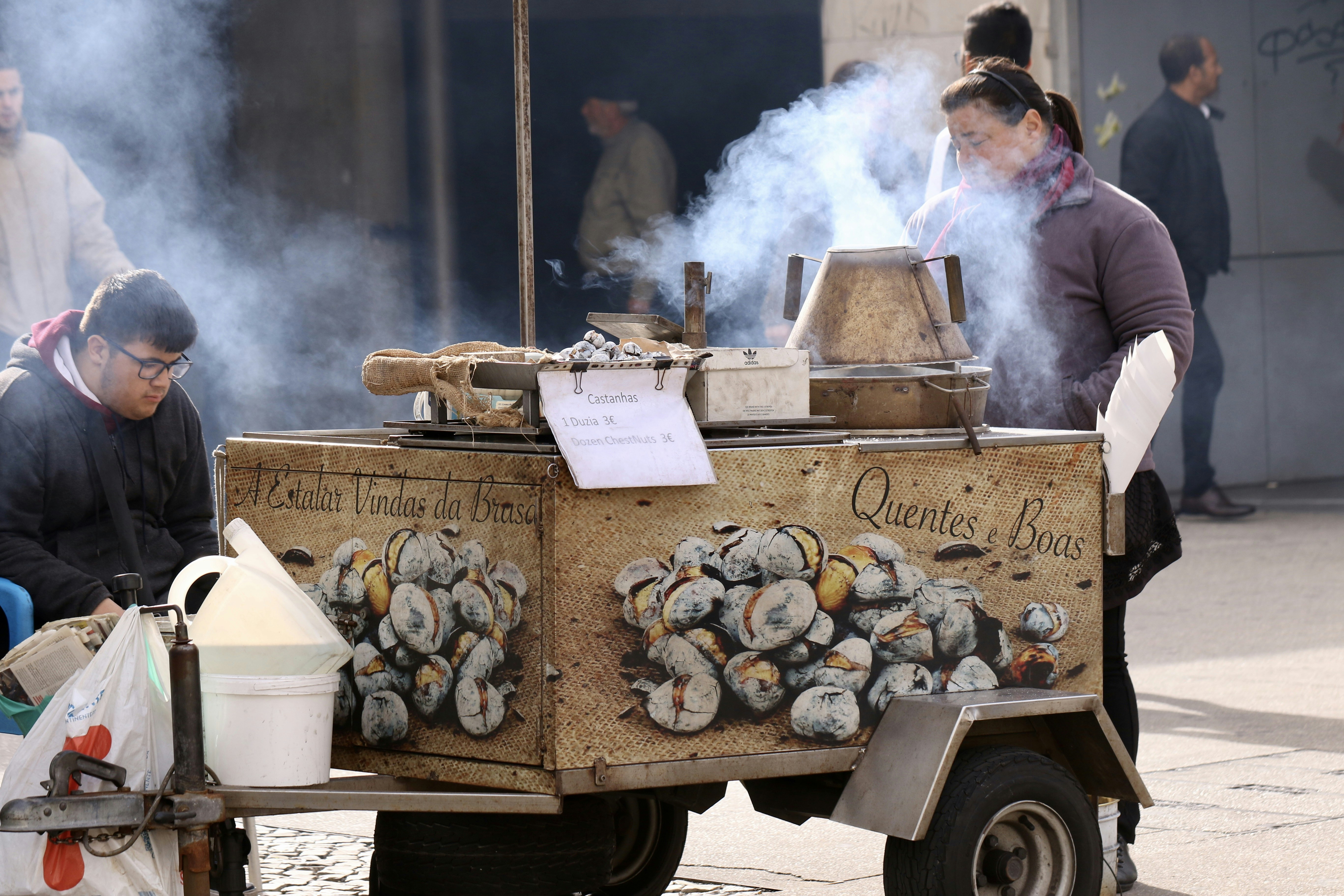 Street vendor cart adorned with traditional designs, serving hot snacks amidst a cloud of steam. A customer enjoys the atmosphere.