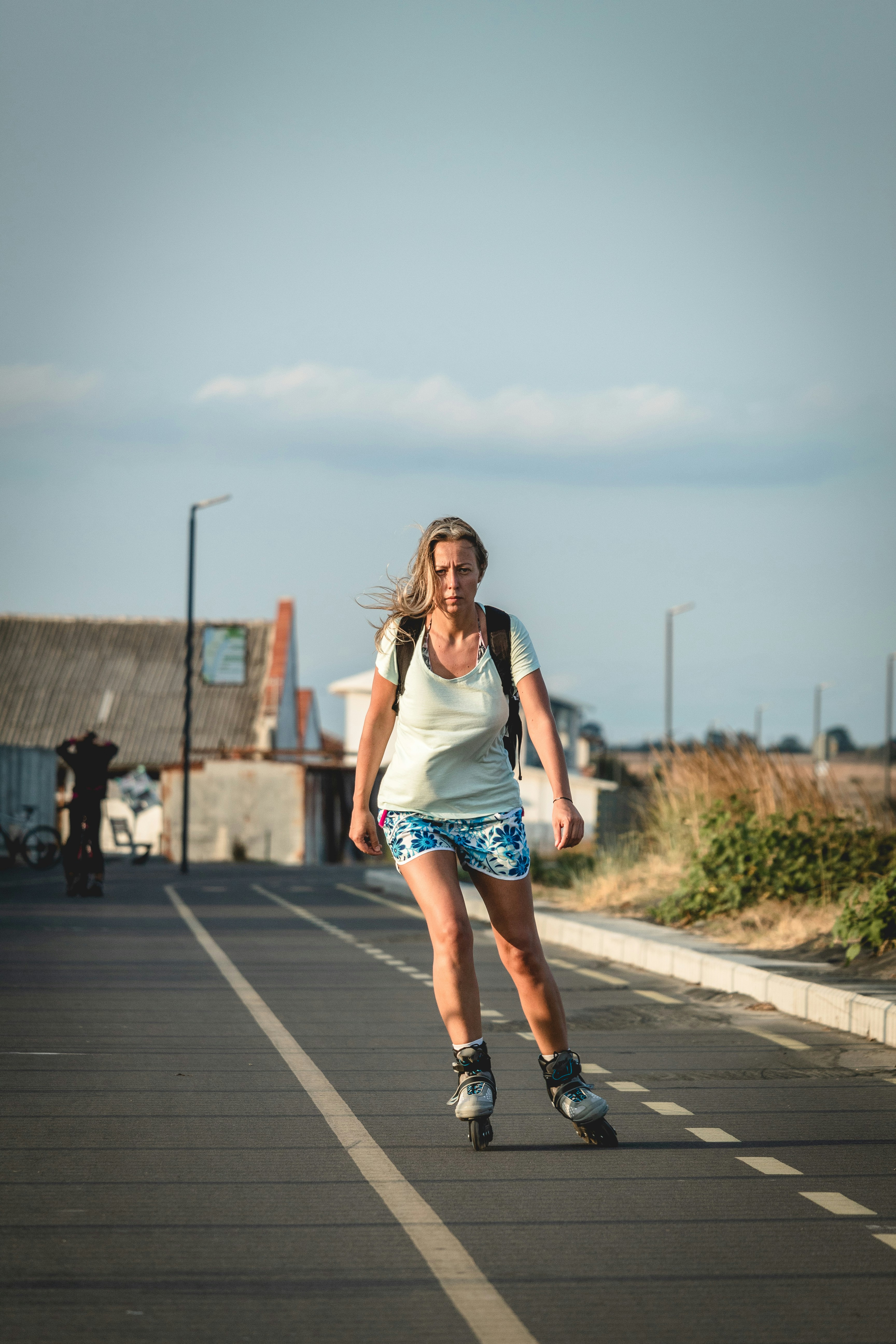 Photograph of a woman rollerblading along a coastal promenade in daylight, wearing a white top, blue floral shorts, and a backpack.
