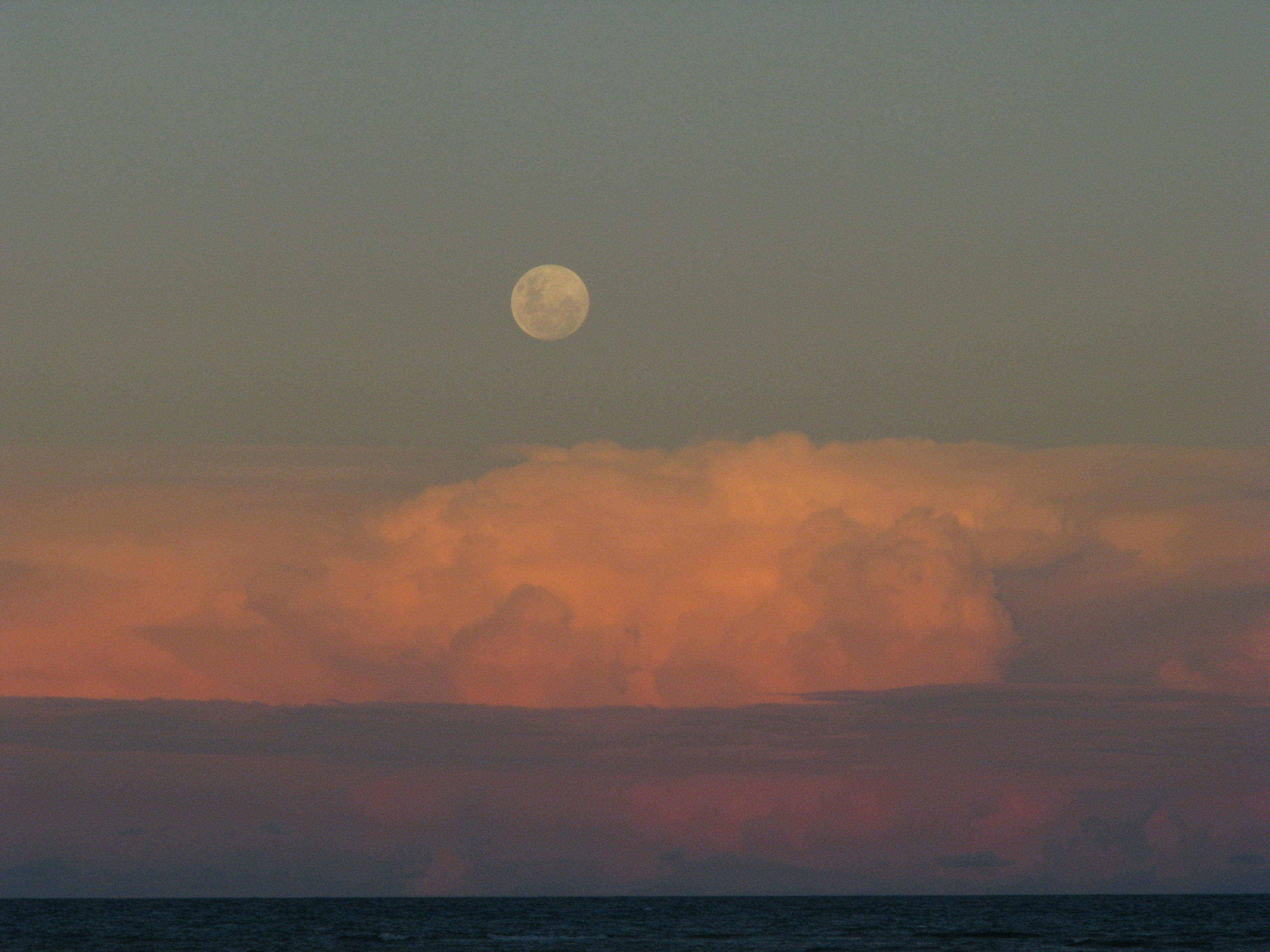 Full moon rising above vibrant, pink-hued clouds at dusk.