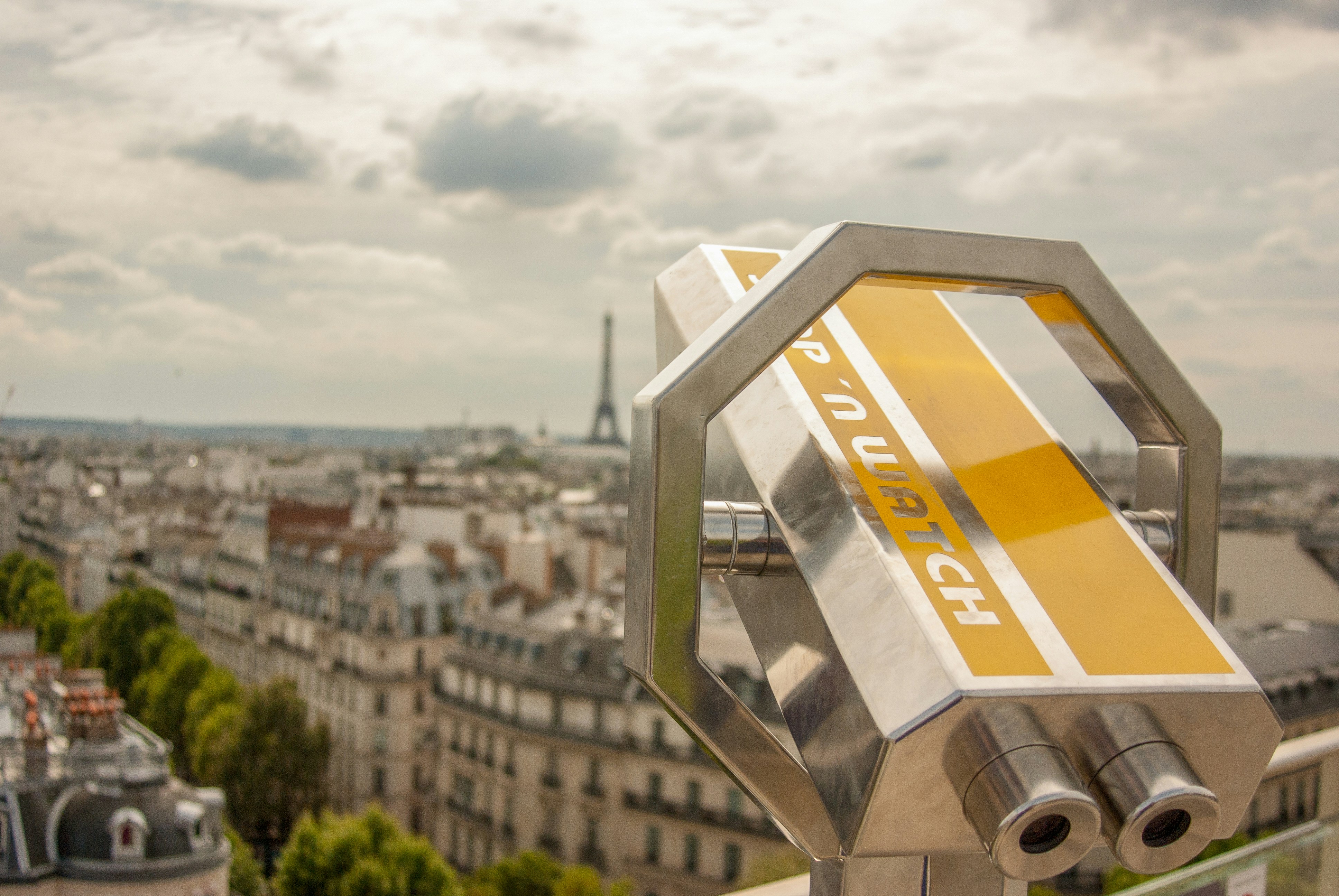 Close-up photograph of a metallic coin-operated viewing device in the foreground, set against a Paris skyline. The Eiffel Tower peeks through the cityscape in the distance.