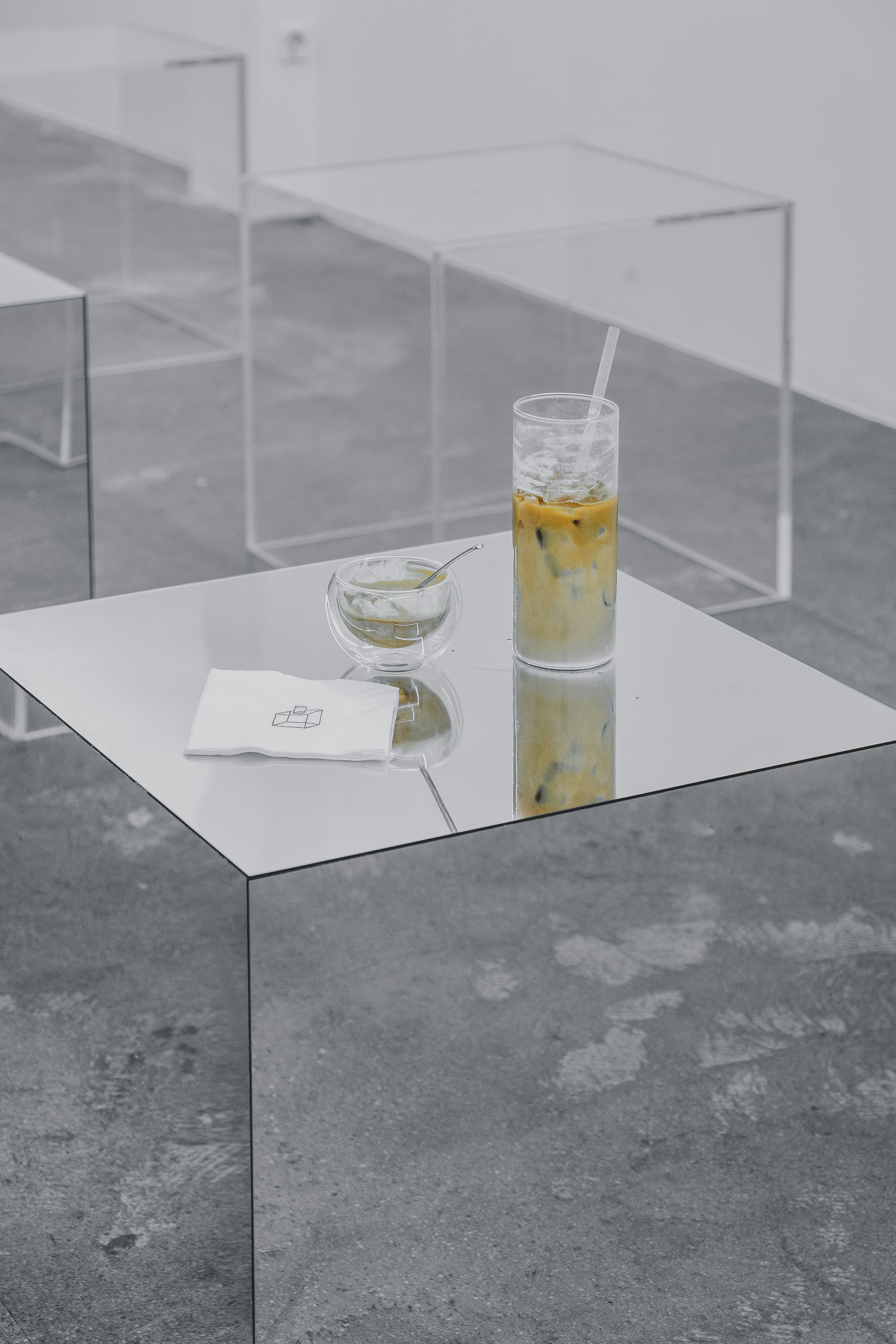 Tall iced beverage on a mirrored table beside a small glass with a spoon and a folded napkin; reflections emphasize the geometric minimalism of the scene.
