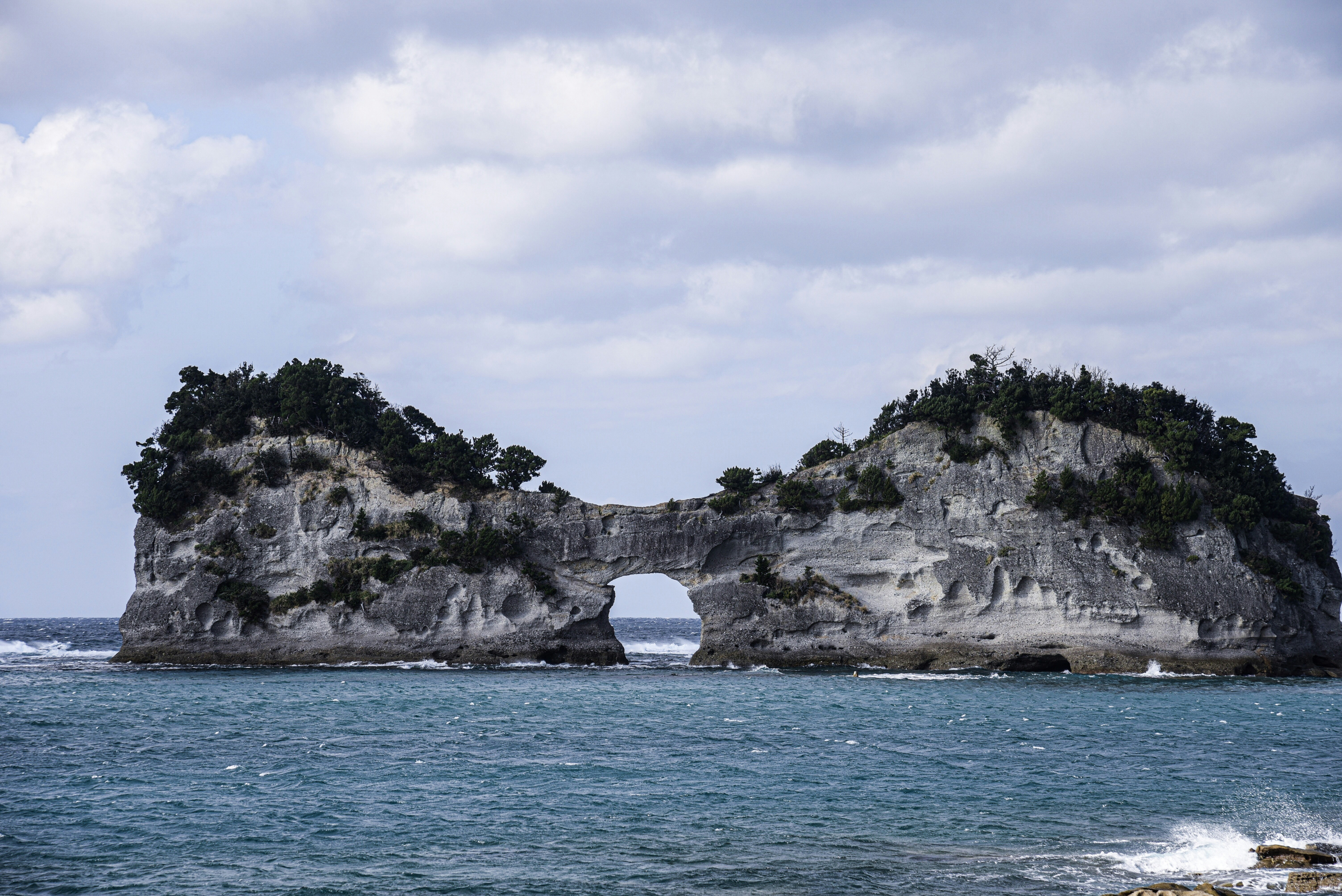 Unique rock formation with a natural arch surrounded by ocean and lush greenery. Ideal for showcasing geological beauty.