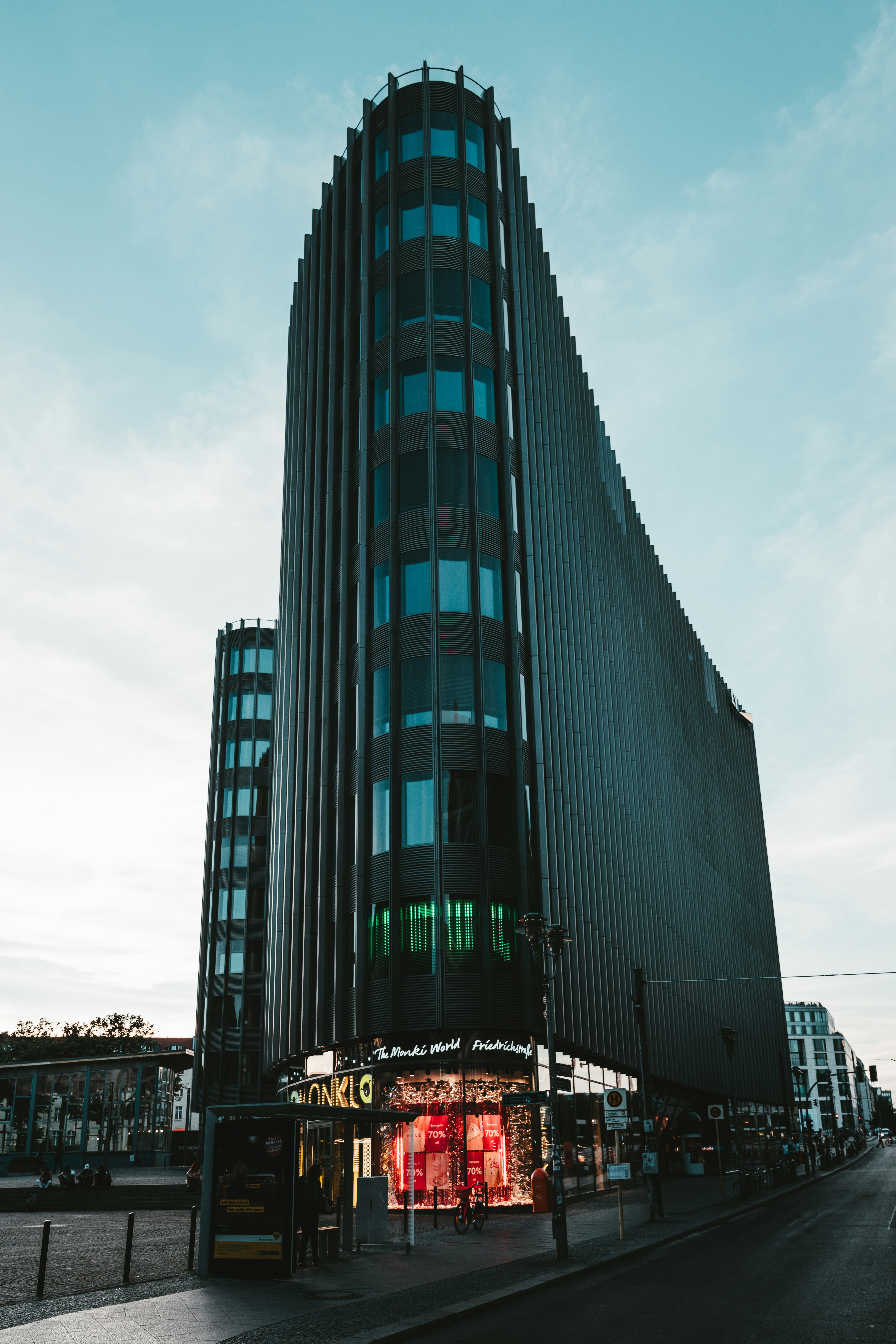 Sleek, contemporary building with a curved facade and large glass windows, reflecting the sky. The ground level features vibrant storefronts and a bus stop.
