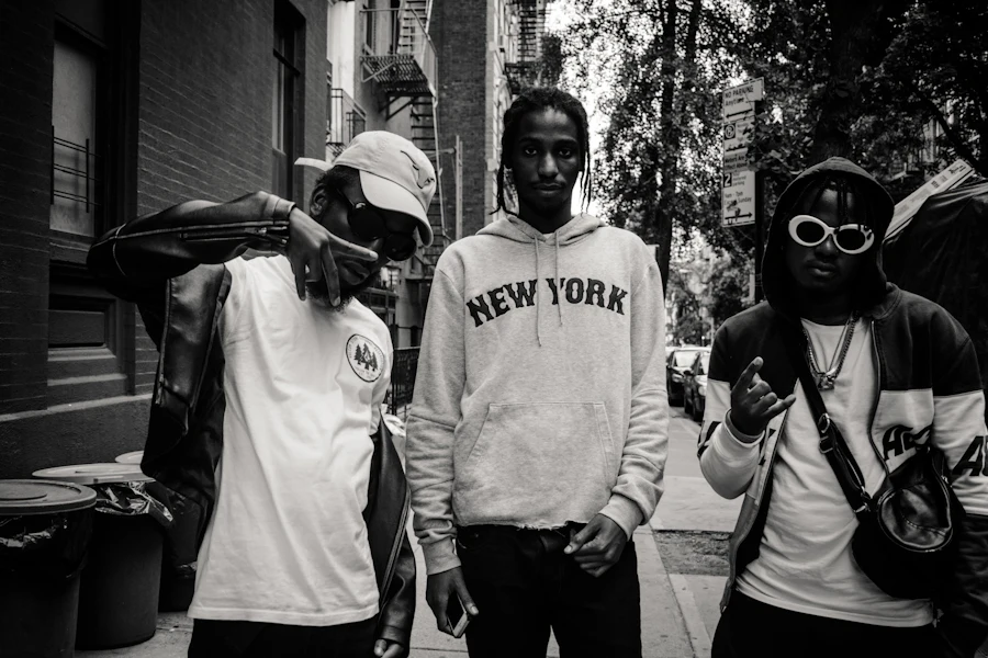 Three young men pose on a narrow urban street, one centered in a “NEW YORK” hoodie and the others wearing streetwear and sunglasses. Black-and-white tones emphasize the gritty city setting and casual attitude.