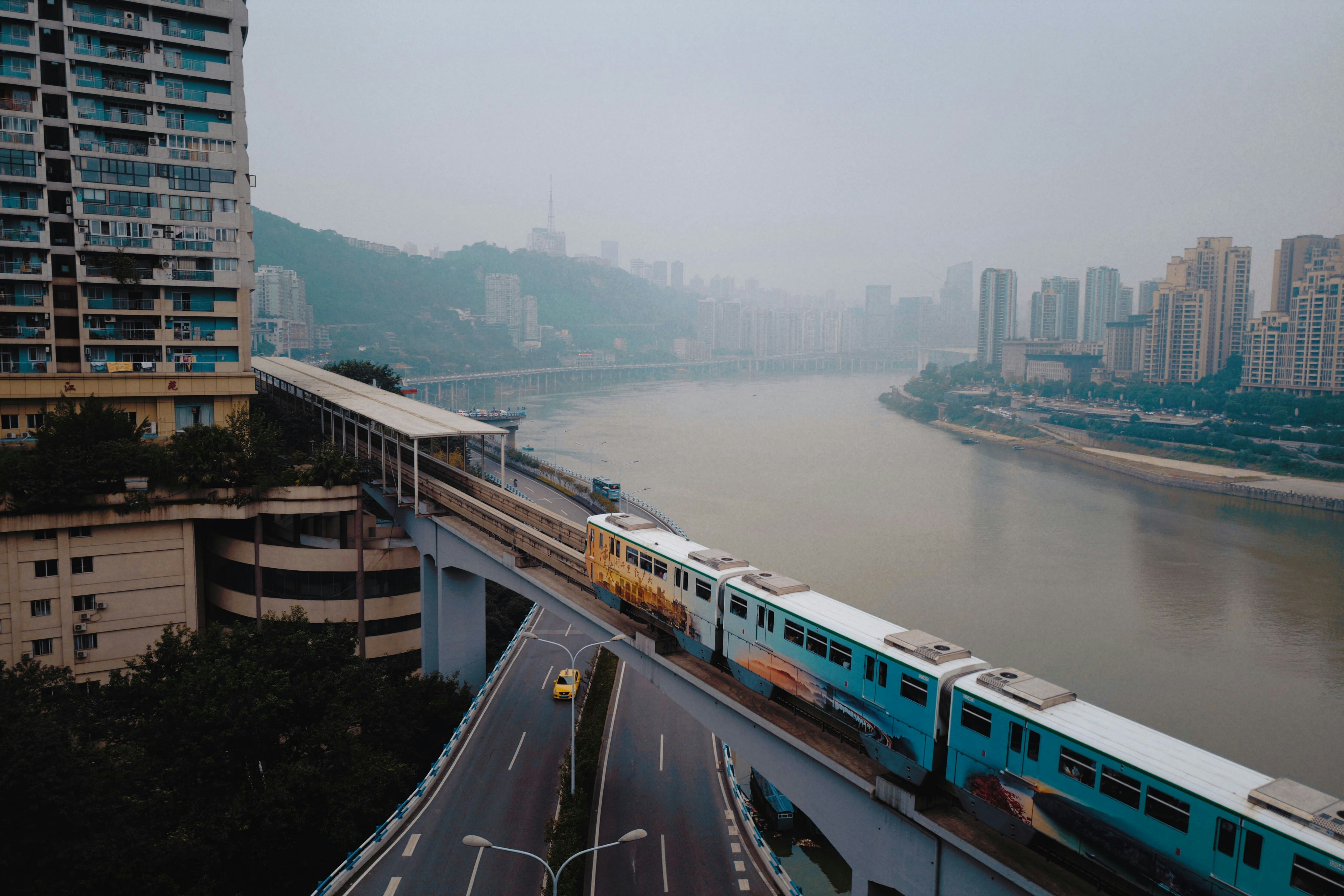 Train crossing a bridge over a wide river with cityscape and misty hills in the background.