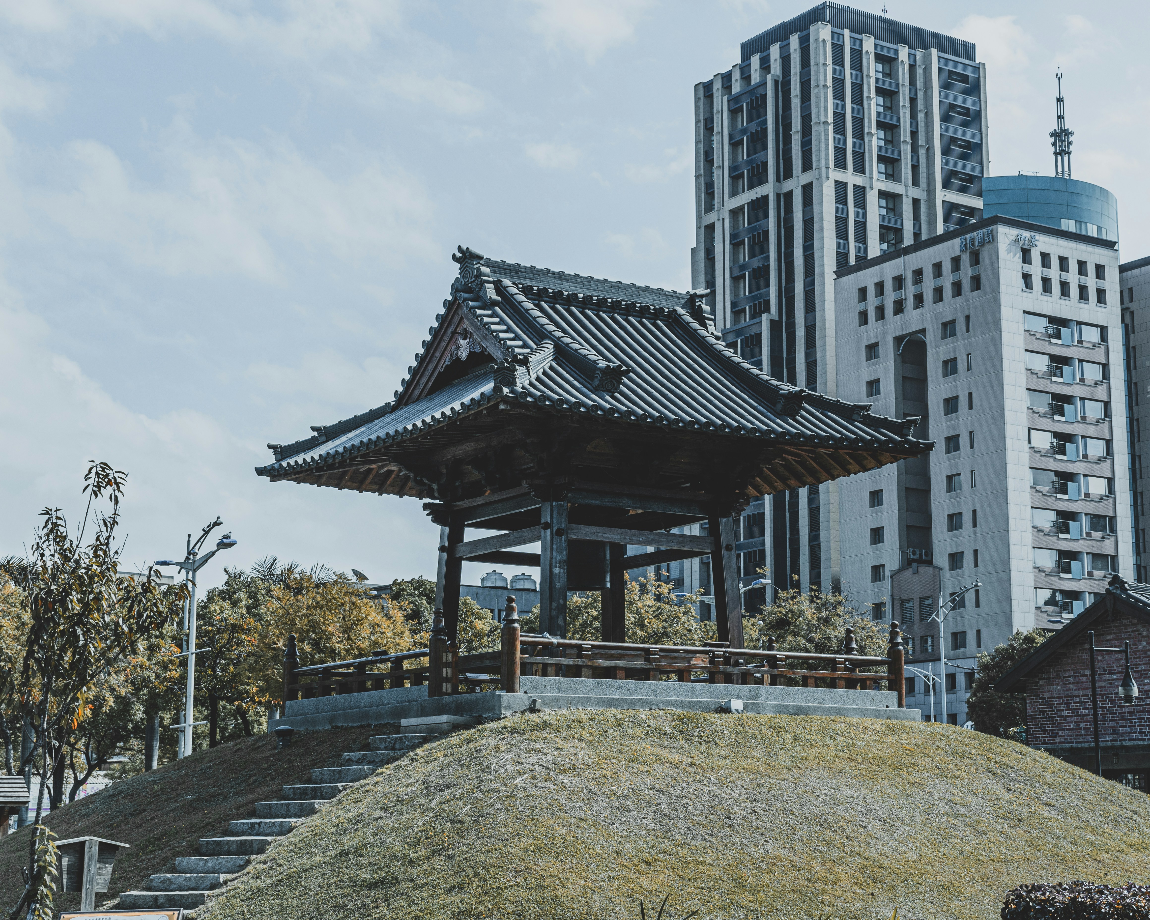 Traditional pavilion set on a grassy mound, surrounded by modern skyscrapers and trees in an urban park.