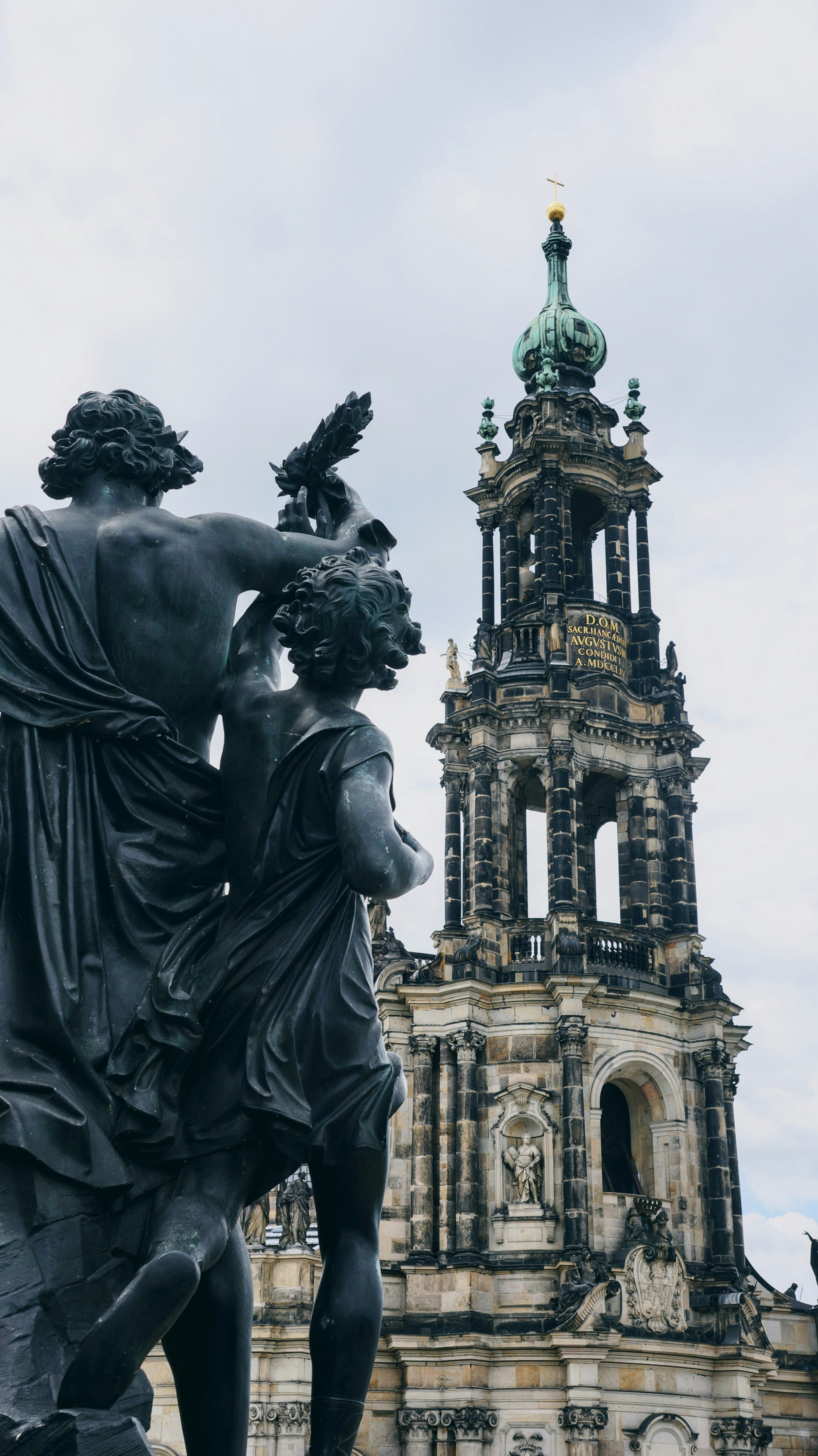 Photograph capturing a bronze statue pair in the foreground with a Baroque bell tower rising behind, under a pale cloudy sky.