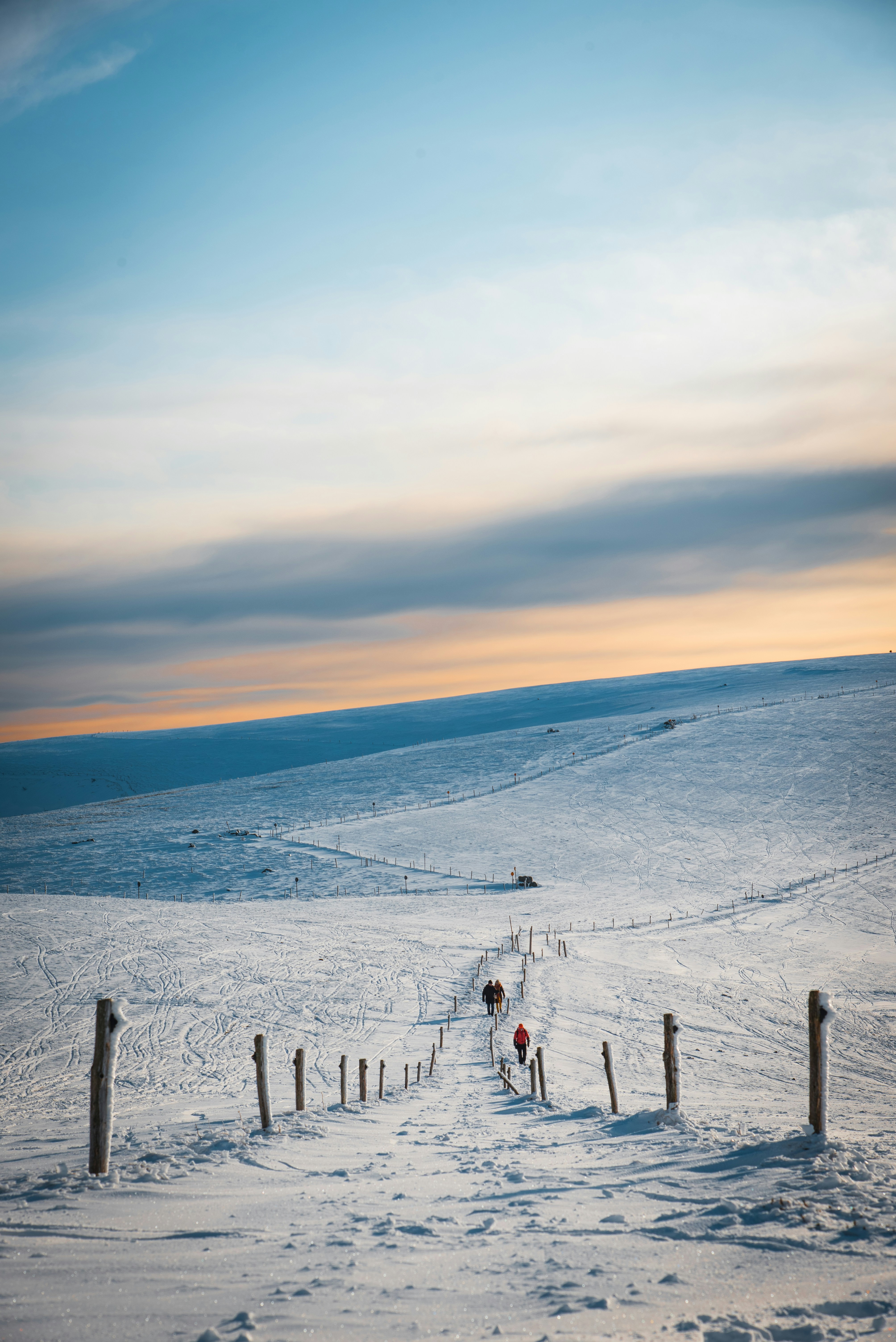 Fence-Lined Snow Trail at Dusk: Two hikers trace a quiet ridge toward the horizon
