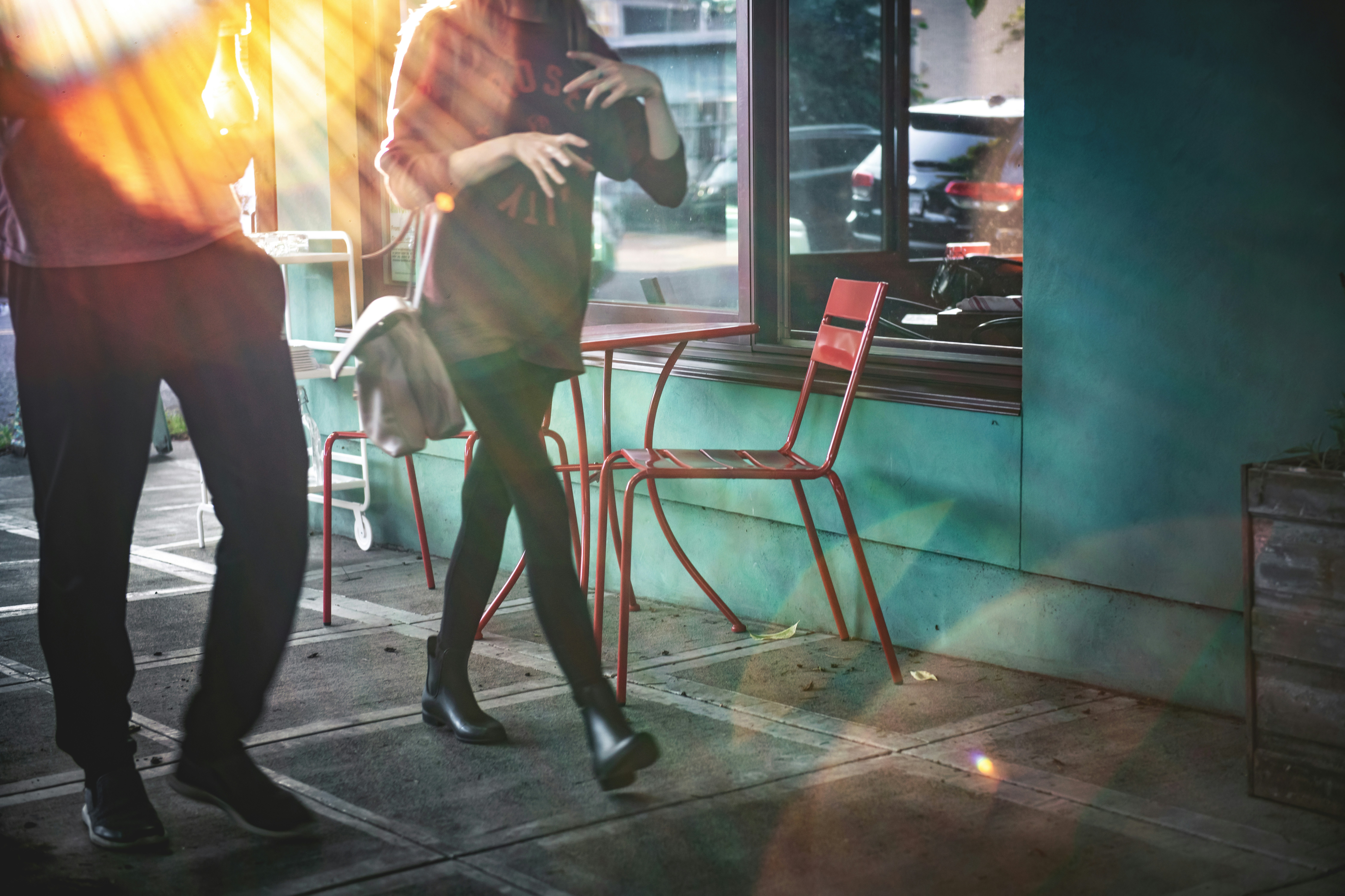 Two people walking past a teal wall with red chairs, illuminated by streaks of sunlight.