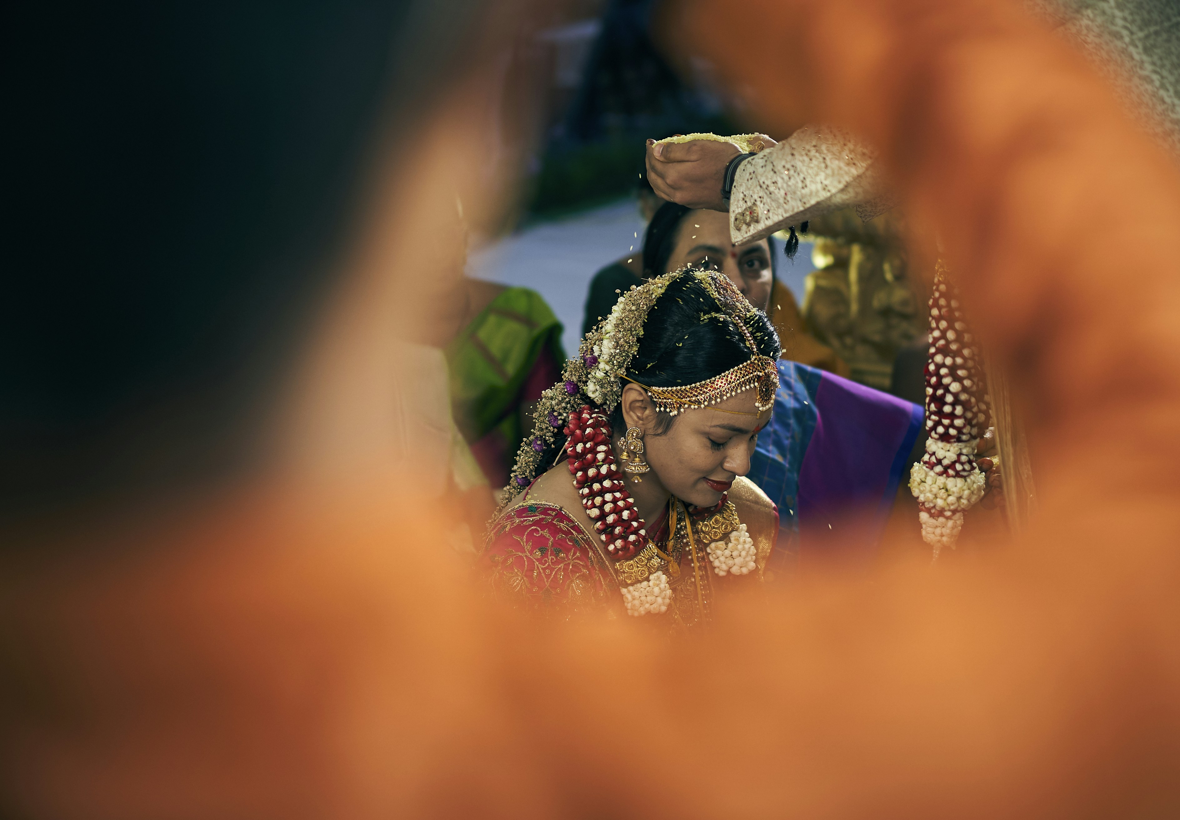 Bride adorned in traditional attire viewed through a garland, highlighting cultural ceremony.