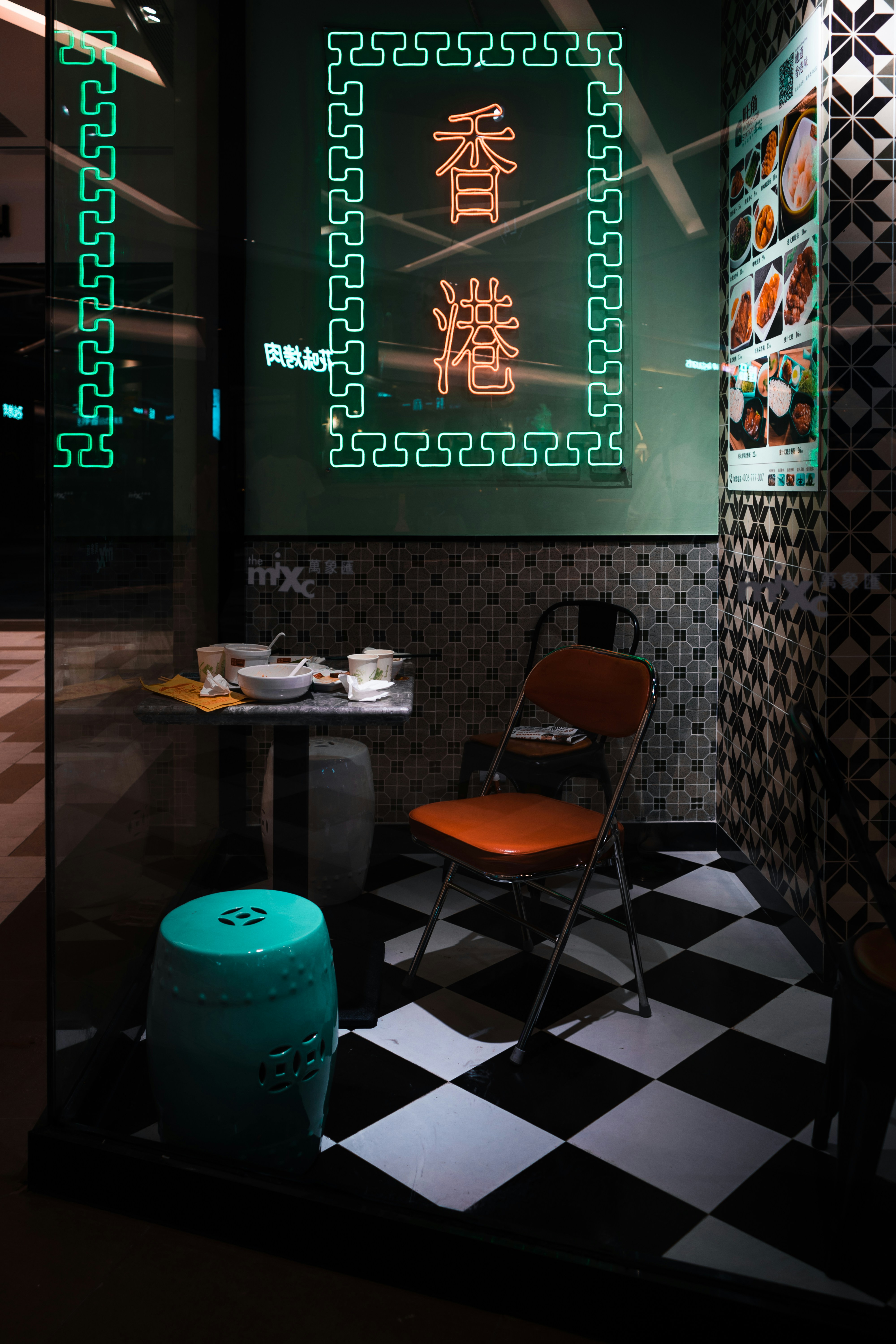 A cozy corner of a restaurant featuring a neon sign, a folding chair, and a turquoise stool, set against a patterned floor. The ambiance suggests a blend of modern design and traditional elements.