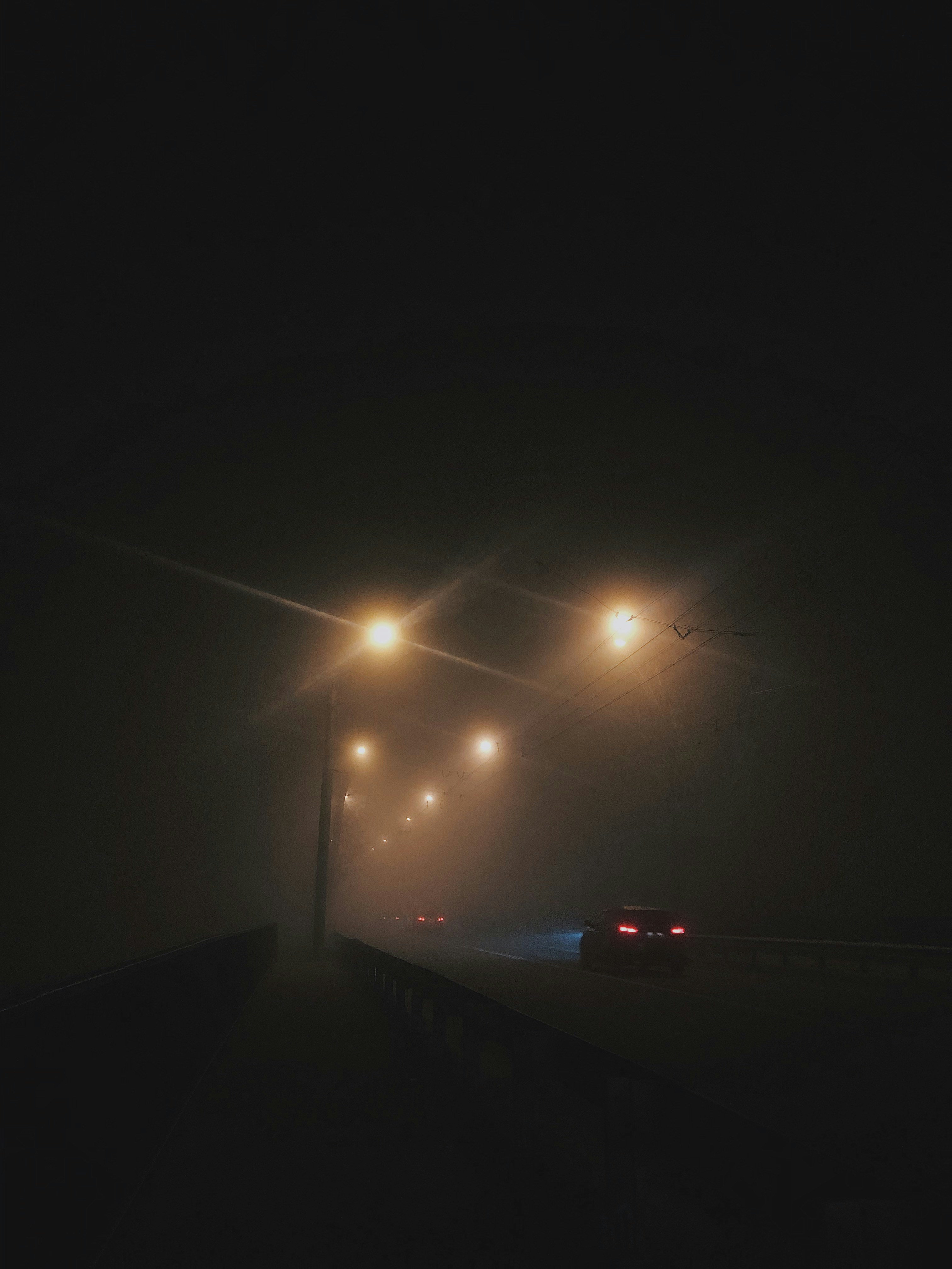 Dimly lit road shrouded in fog, with glowing streetlights illuminating the path ahead. Two cars are visible in the distance, adding depth to the scene.