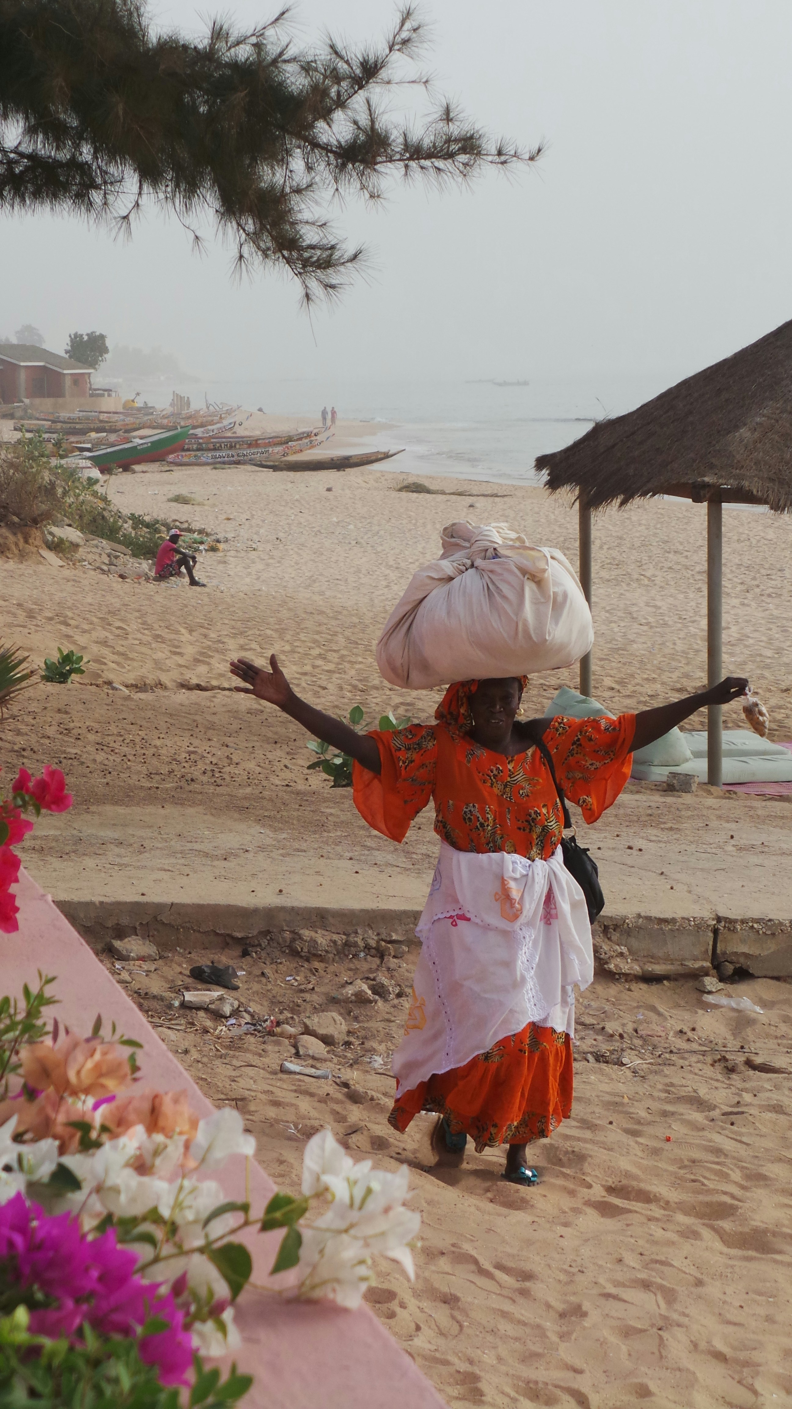 A woman in vibrant attire joyfully carries a large bundle on her head along a sandy beach, with fishing boats in the background and colorful flowers in the foreground.