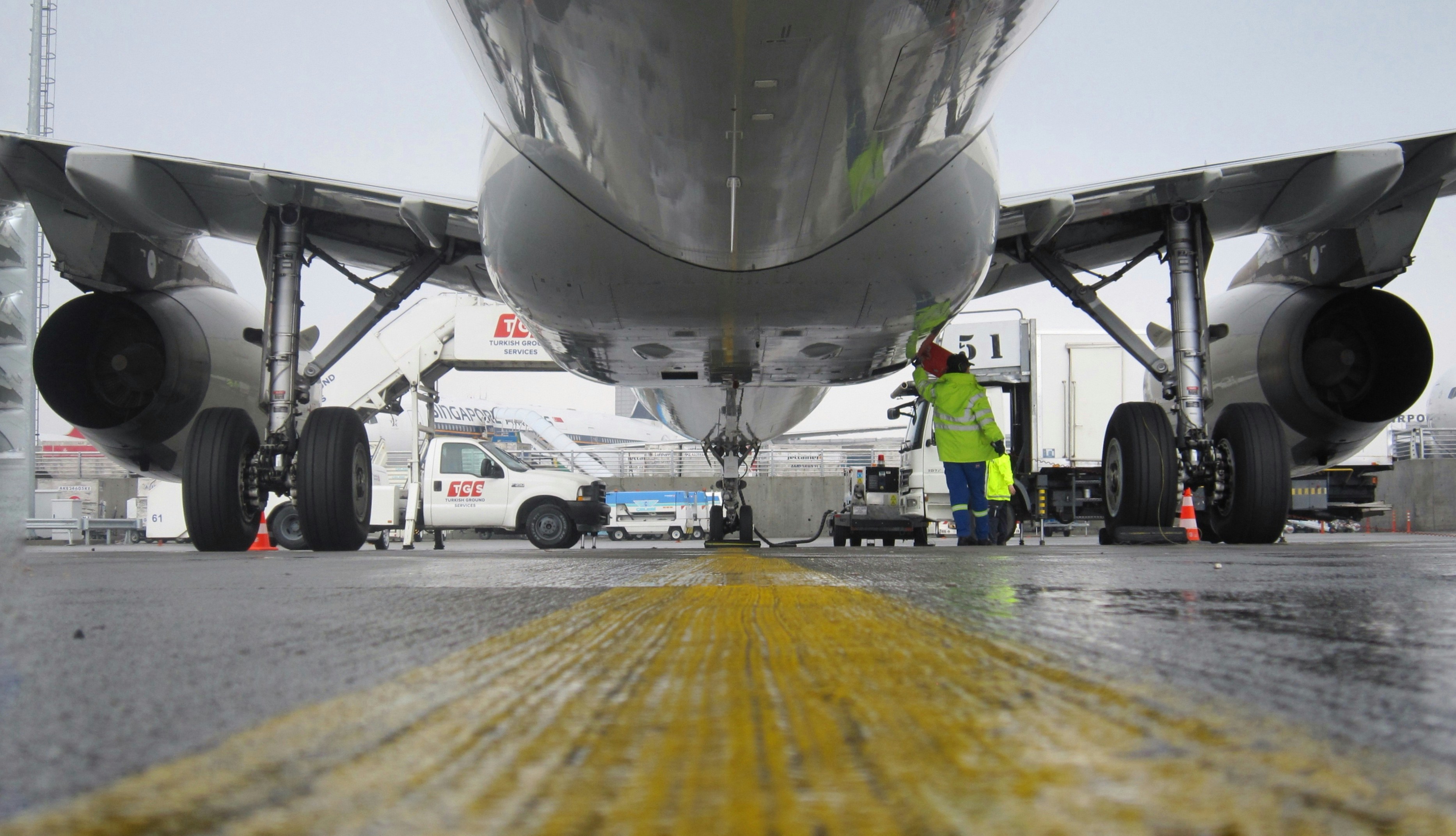 Ground crew member inspecting an aircraft from below while service vehicles are stationed nearby. The focus is on the aircraft's landing gear and engines.