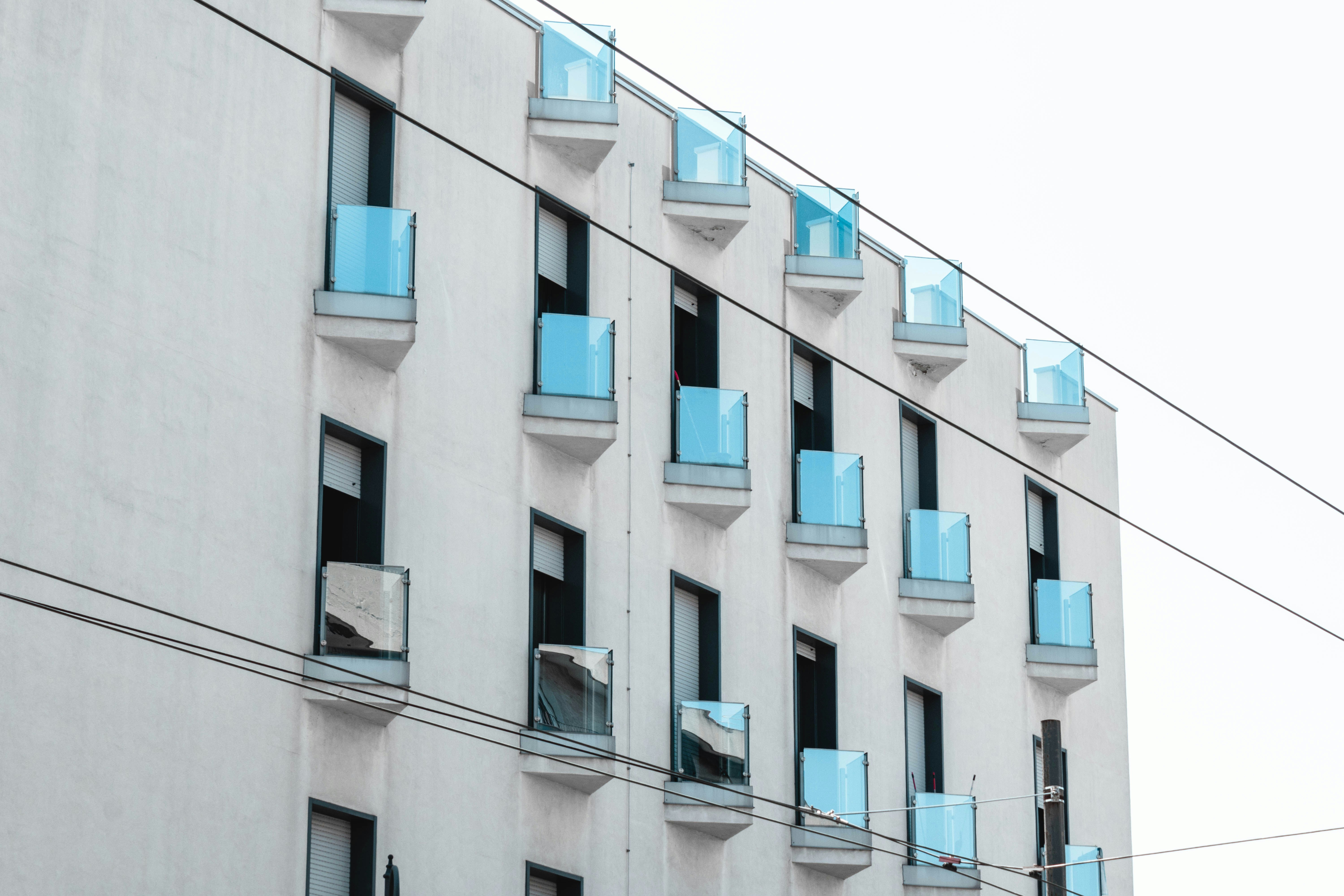 Contemporary building featuring multiple balconies with blue glass railings, set against a pale backdrop.