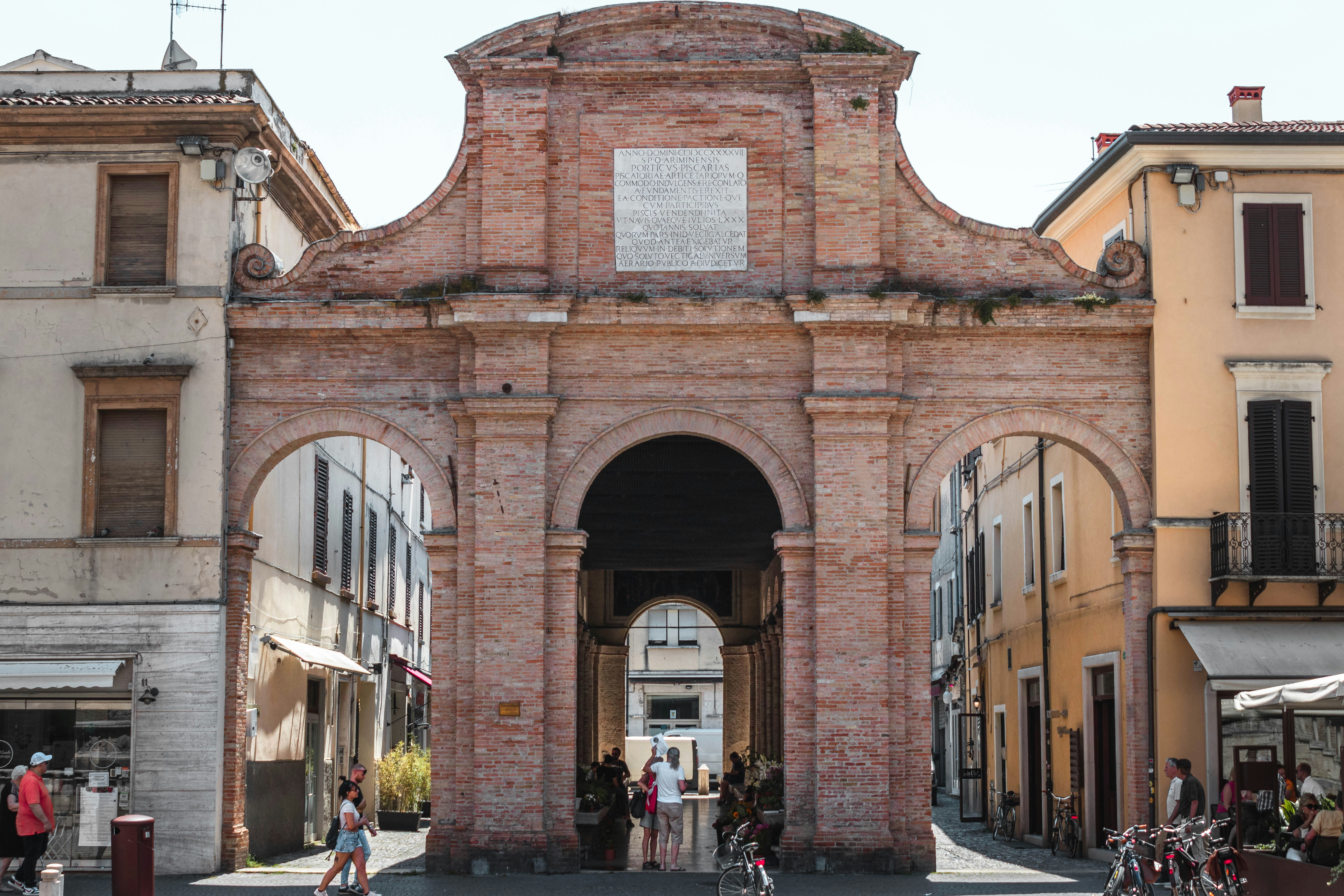 Brick archway flanked by buildings on a bright street, with people passing beneath.