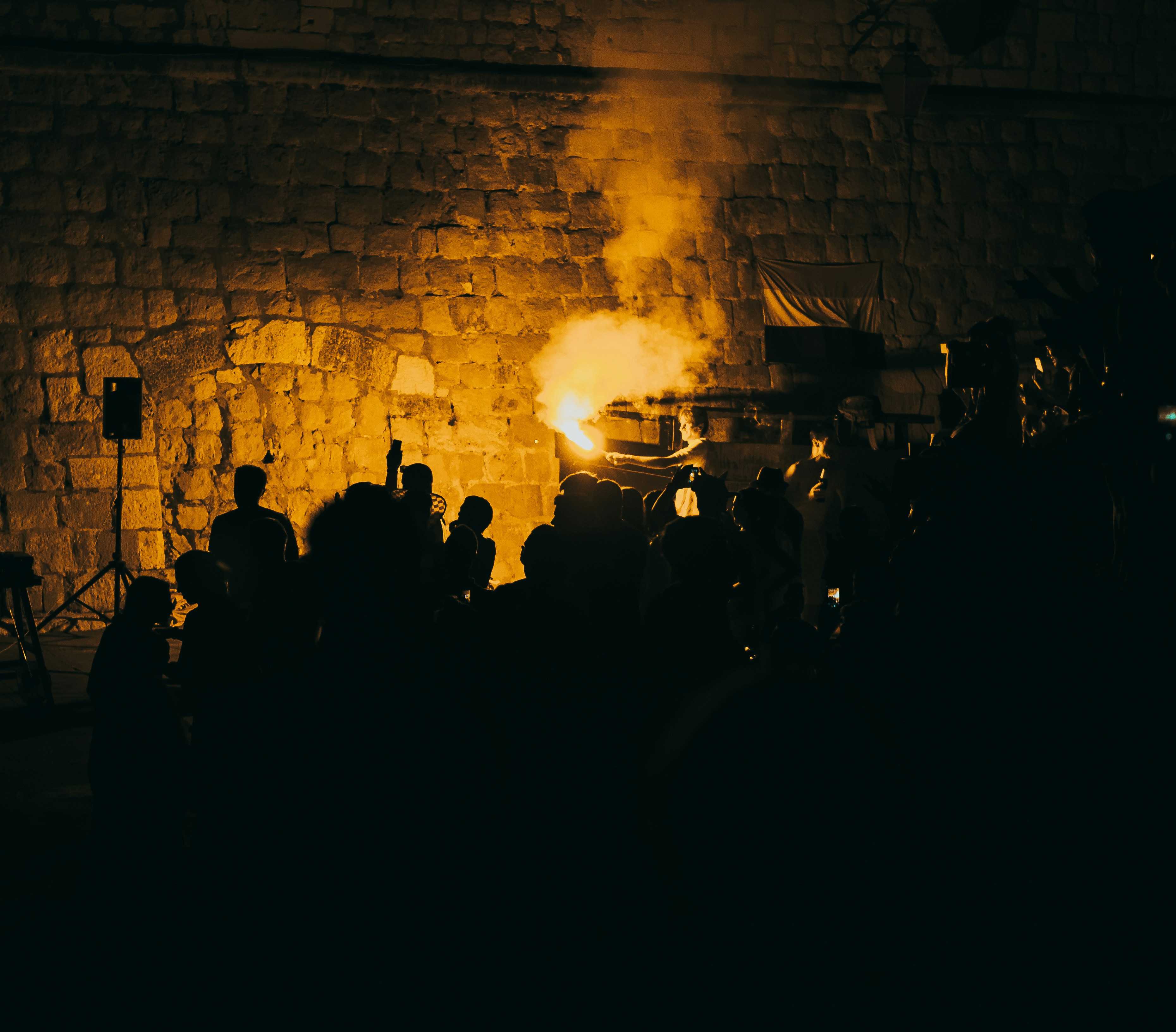 Silhouetted figures gather around a vibrant flame against a textured stone wall, creating a dramatic interplay of light and shadow.