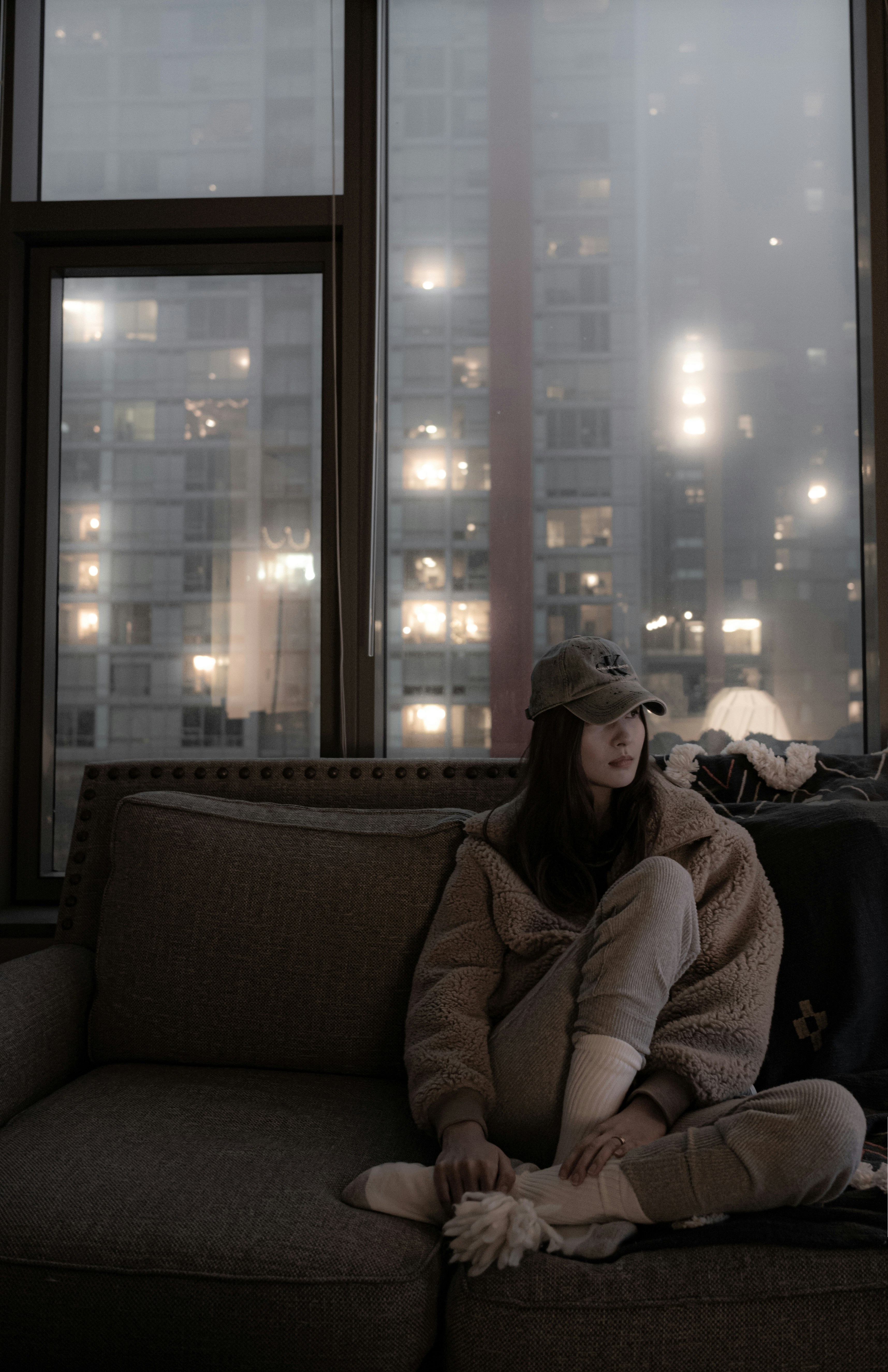 A woman in a fuzzy coat and cap sits cross-legged on a sofa beside a large window, with the city’s lights softly glowing outside.