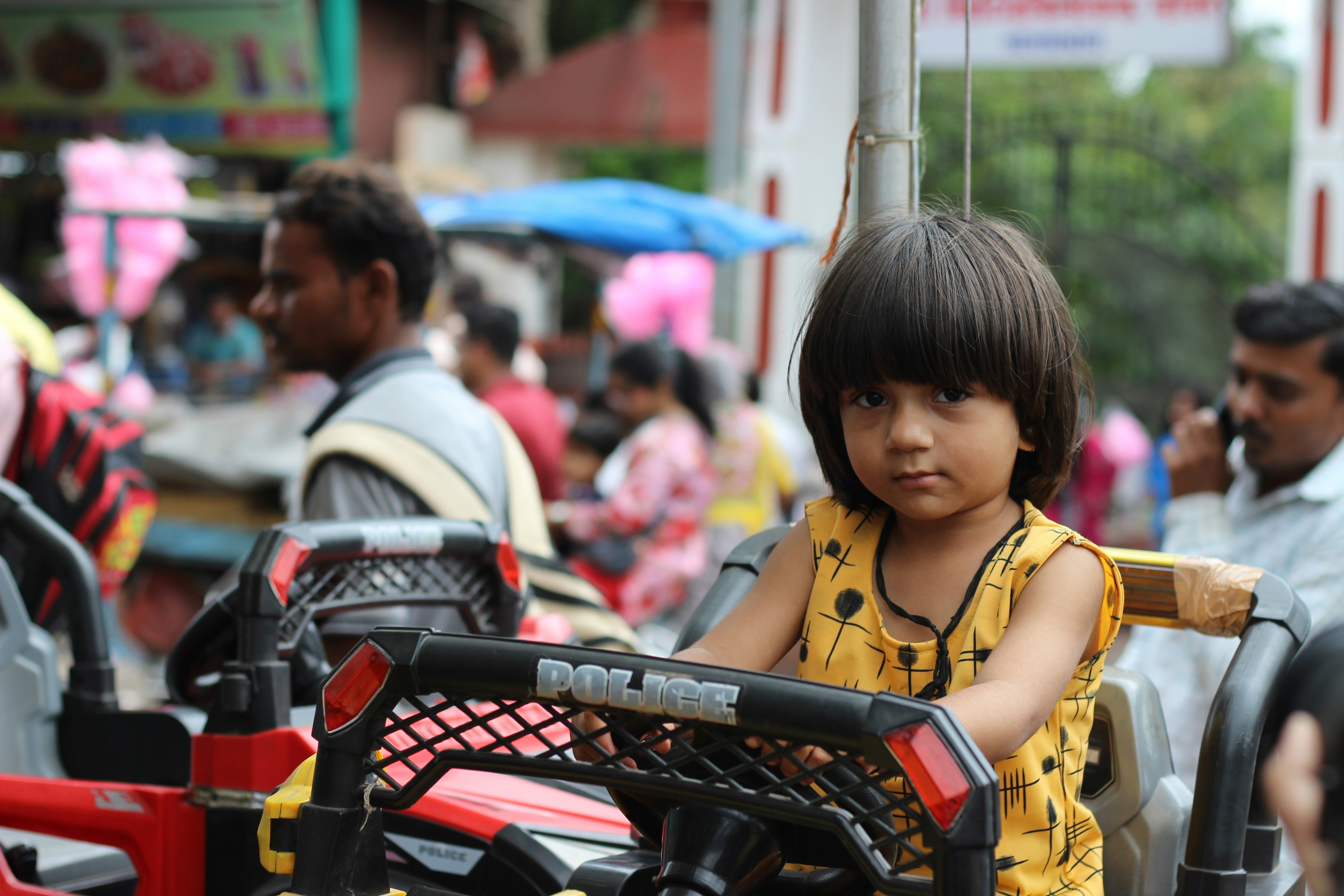 A young child with a playful expression sits in a toy vehicle, surrounded by a bustling fair atmosphere filled with colorful decorations and people.