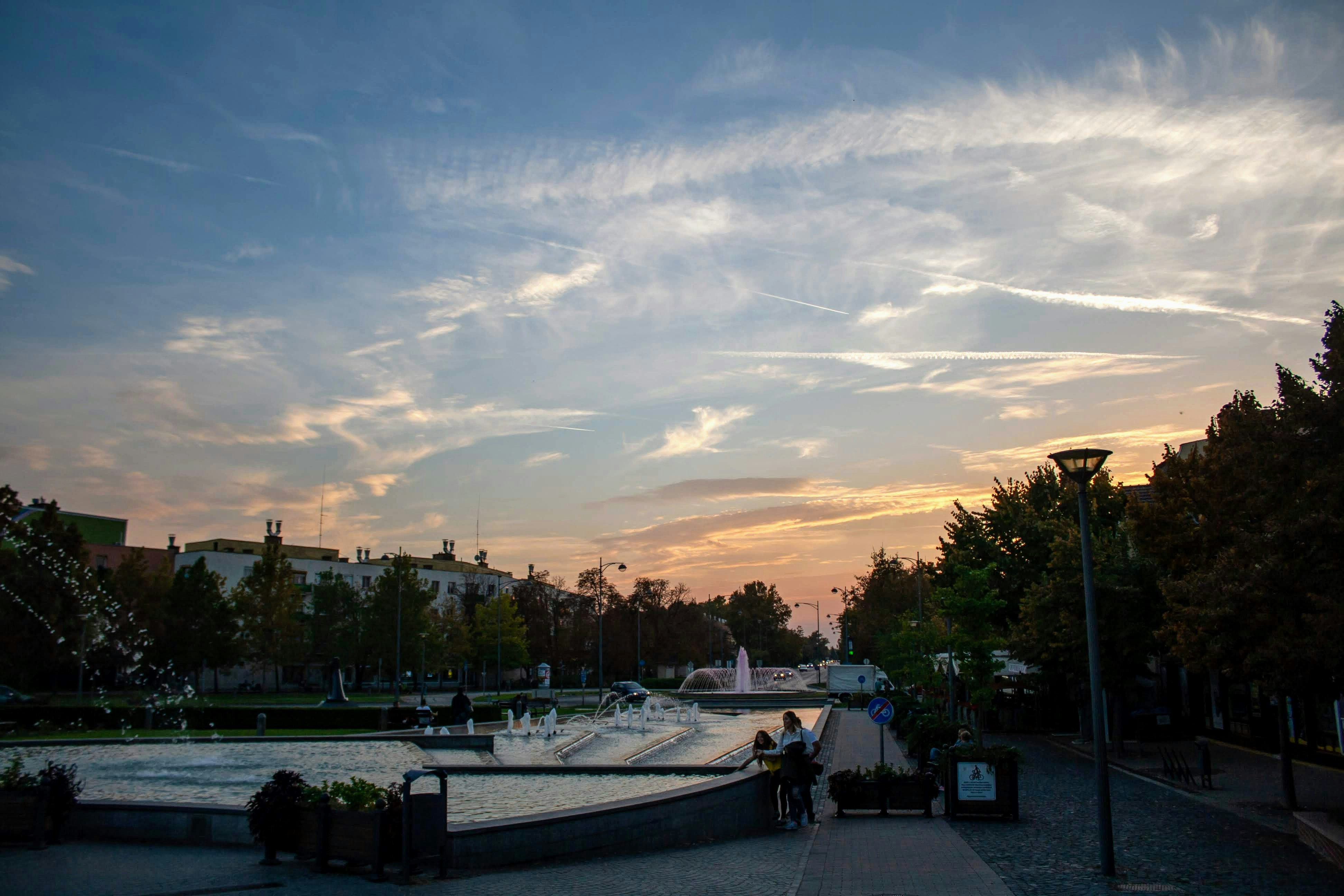 Evening sky over a city park with a fountain and silhouetted trees.