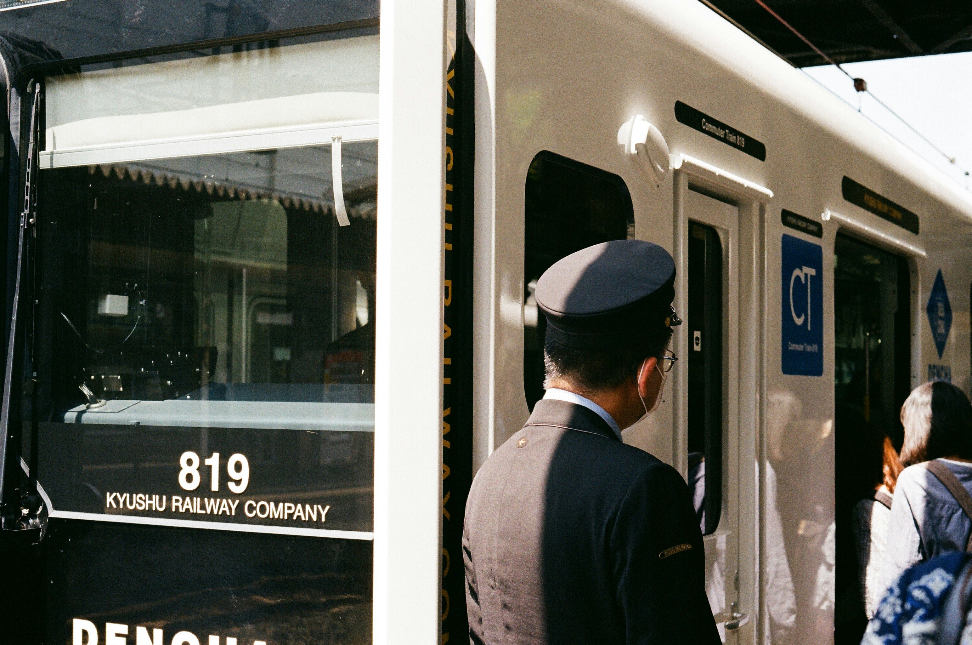 Photograph of a uniformed conductor at the doorway of a Kyushu Railway Company train as passengers board.
