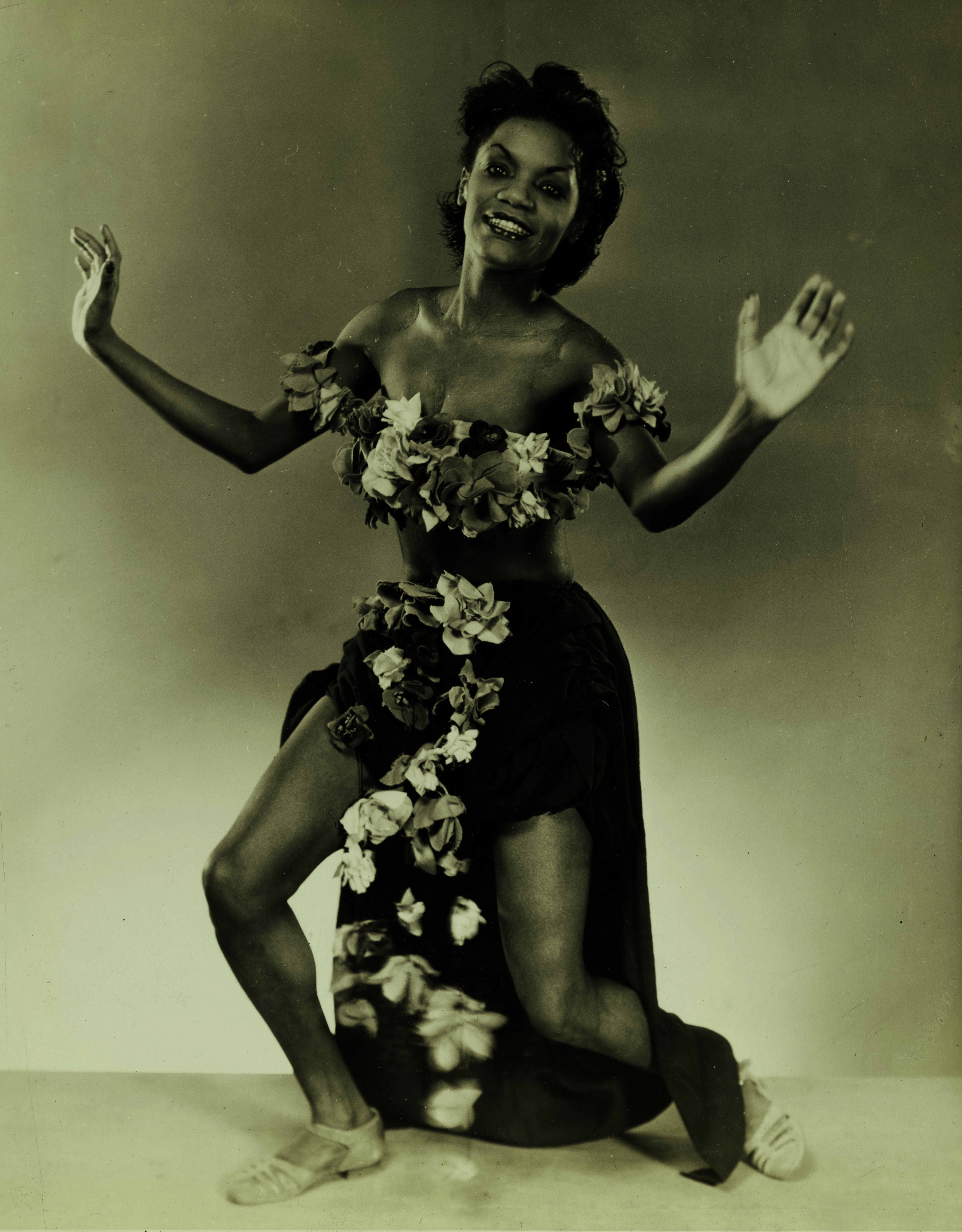 Elegant dancer poses mid-mwing with a garland of flowers; vintage photograph capturing movement and rhythm in a studio setting.