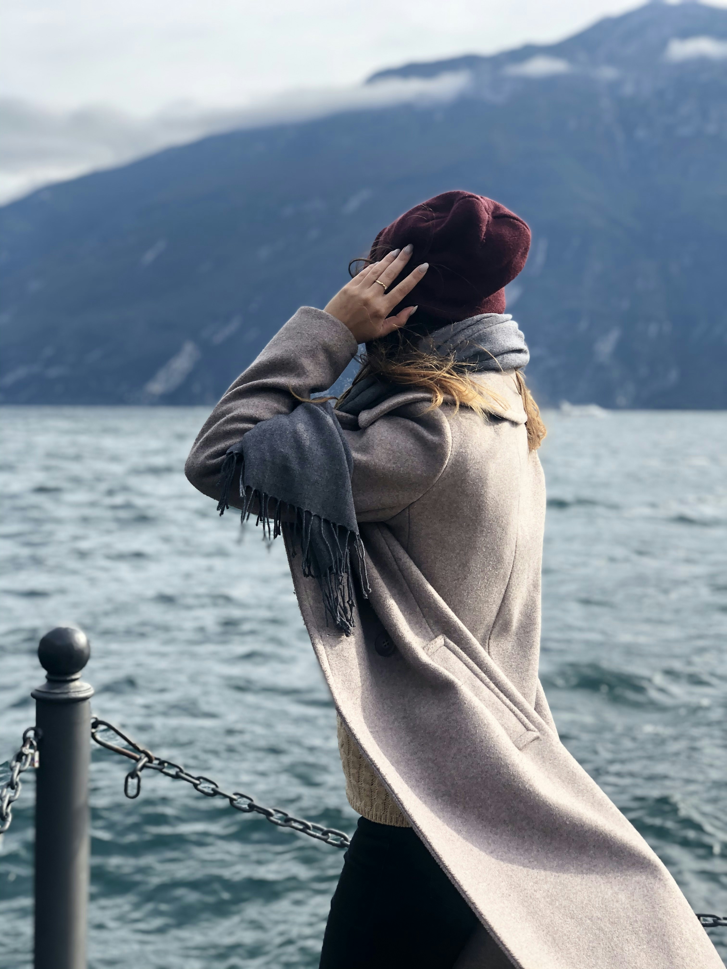 Woman gazing at the serene lake, her coat and scarf fluttering in the breeze against a backdrop of majestic mountains.