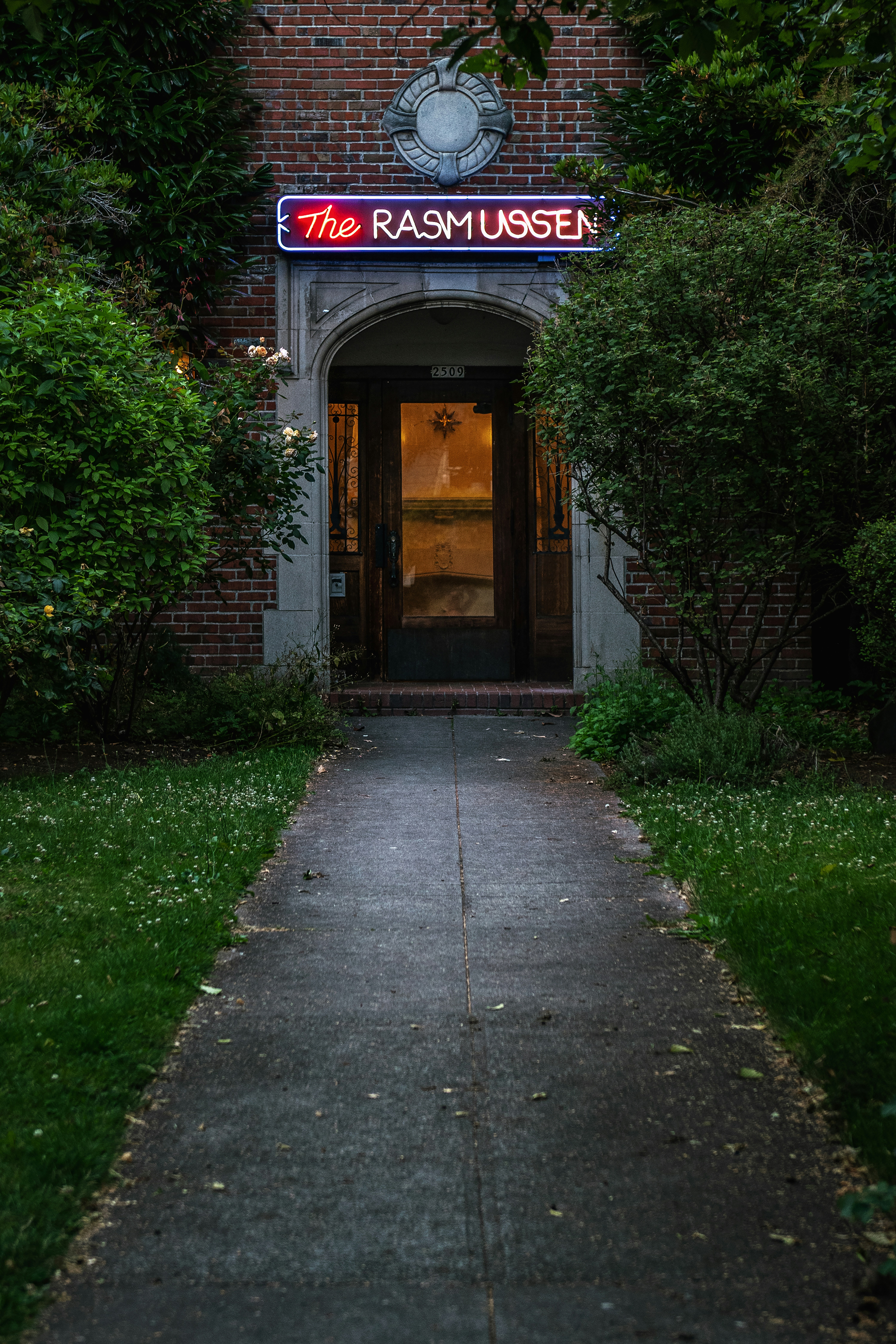 Charming entrance framed by lush greenery and a glowing neon sign, inviting visitors into The Rasmussen.