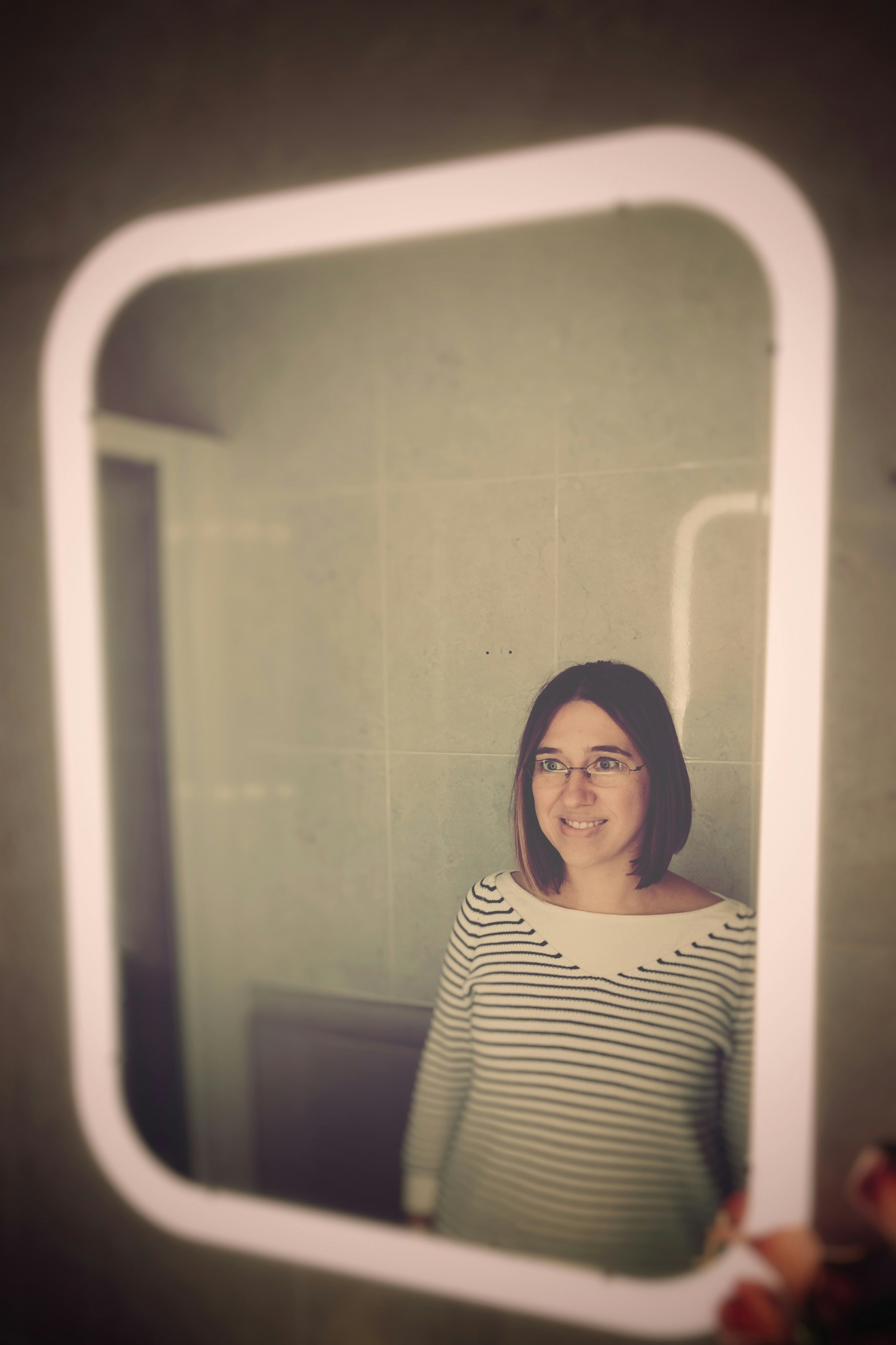 A woman with glasses smiles gently while standing in front of a mirror illuminated by soft light, creating a serene atmosphere.