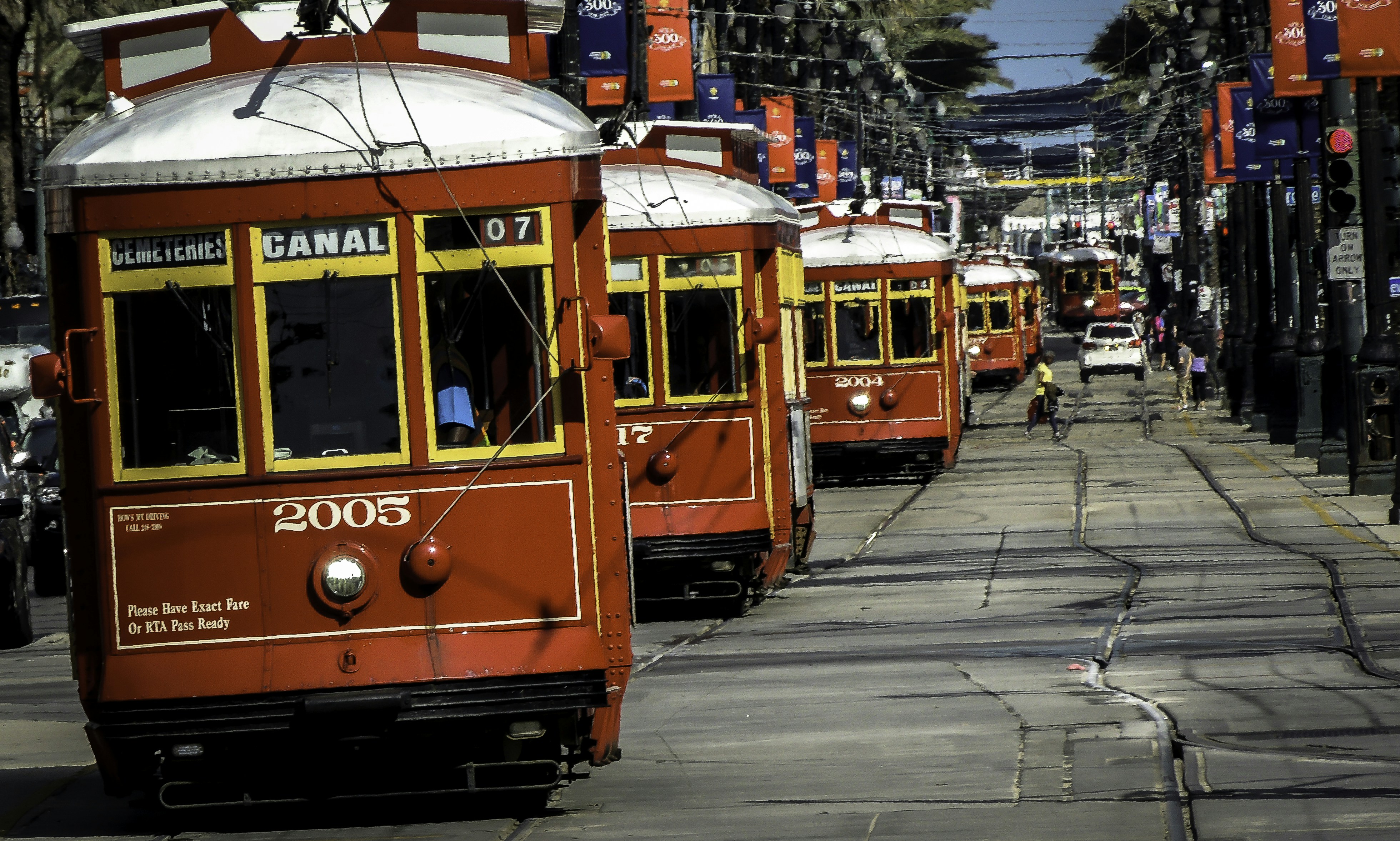 Red trolley cars travel down a street lined with banners and palm trees.