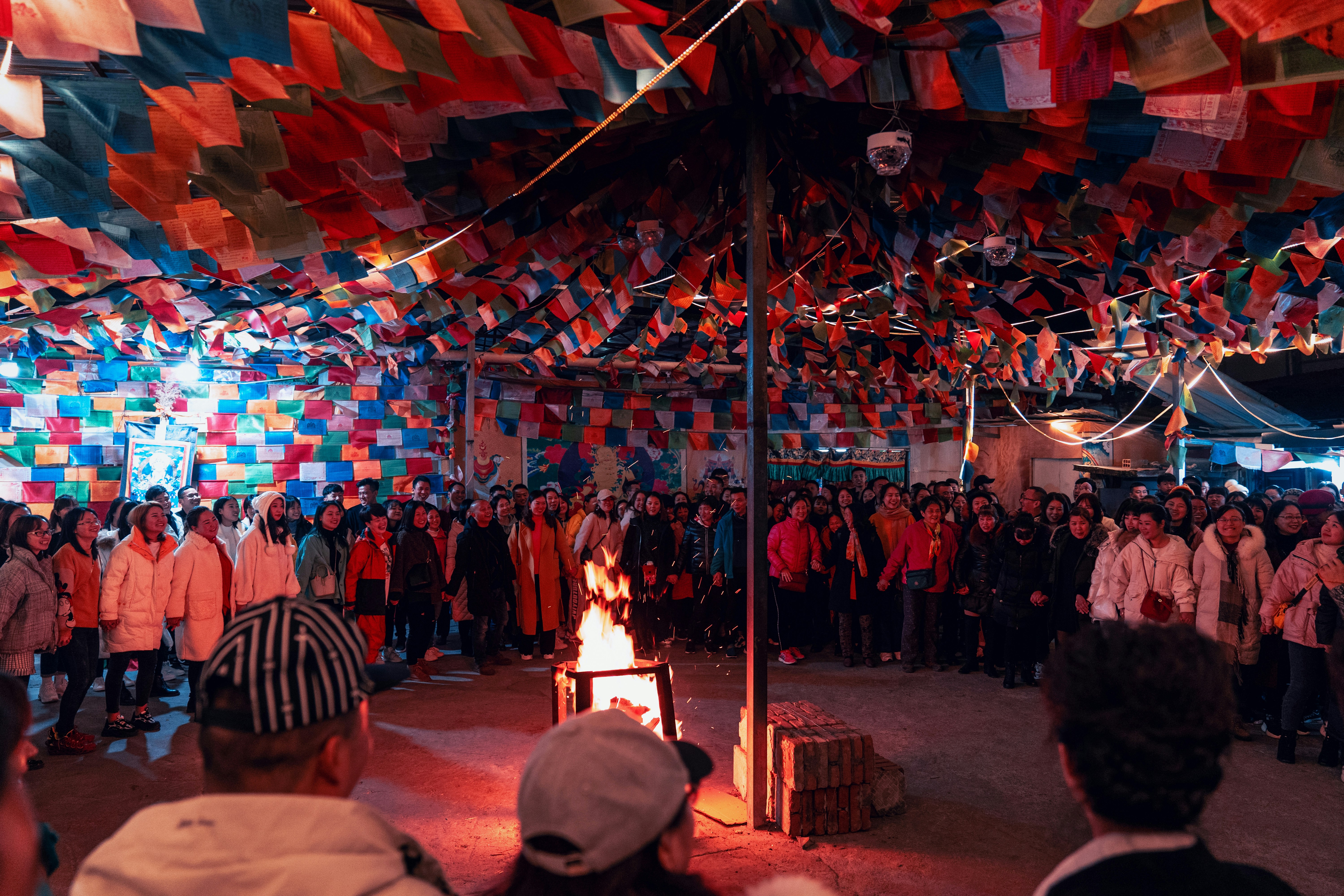 A vibrant gathering of people in warm clothing surrounds a central fire, set against a backdrop of colorful prayer flags. The atmosphere is festive and communal.