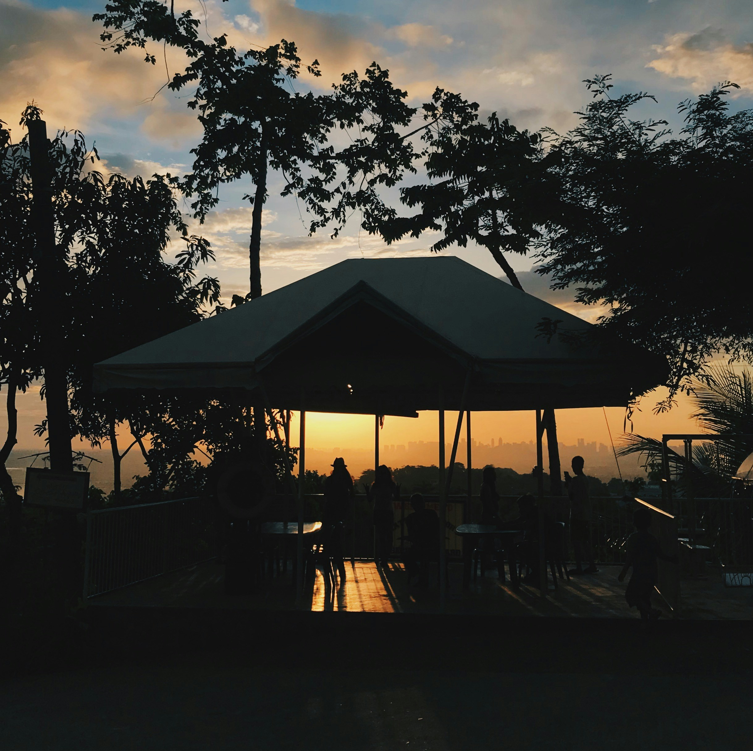 Silhouetted figures gather under a canopy as the sun sets, casting a warm glow over the landscape and creating a tranquil atmosphere.