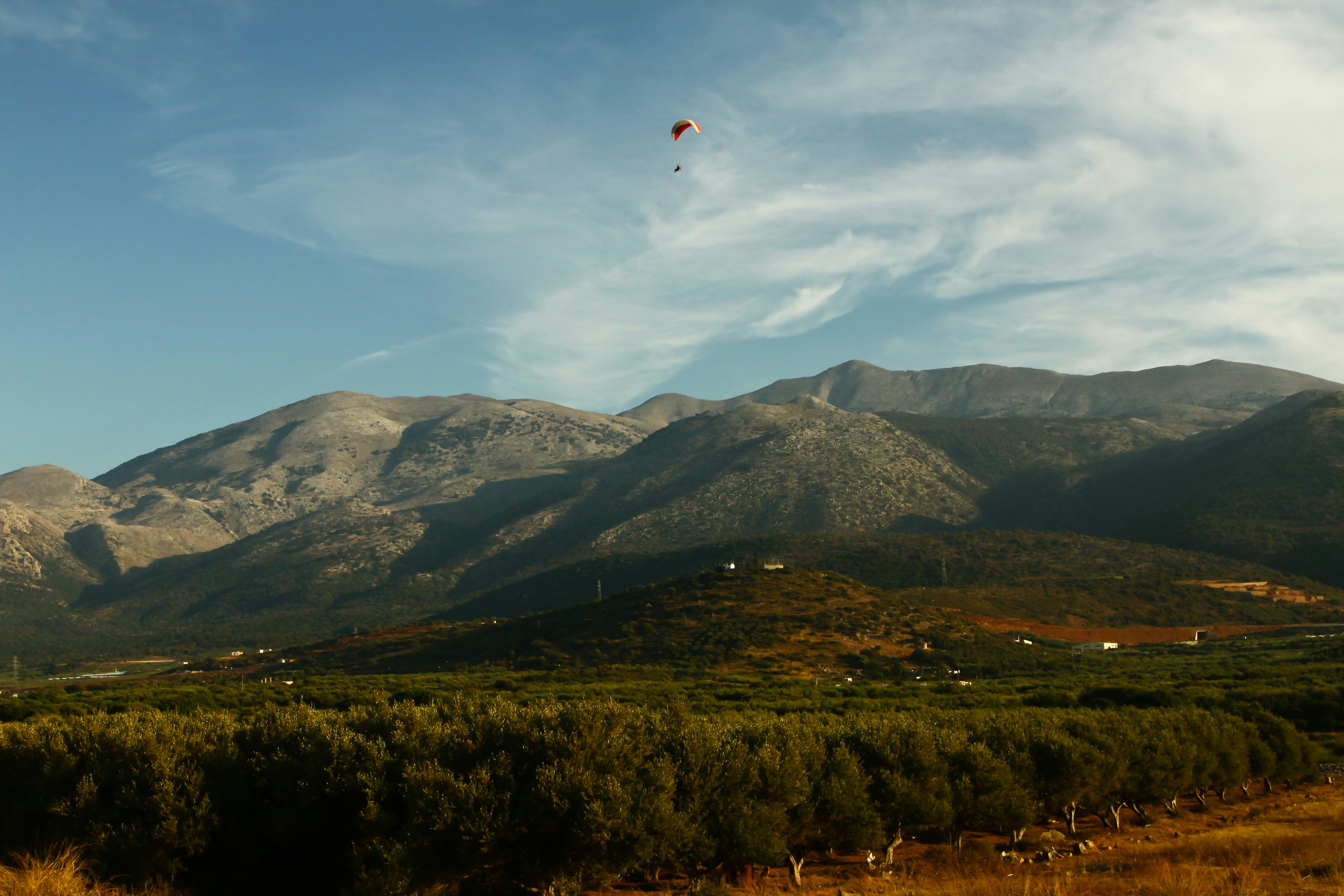 Sunlit valley with olive groves and distant mountain range under a blue sky; a lone paraglider drifts above the ridges.