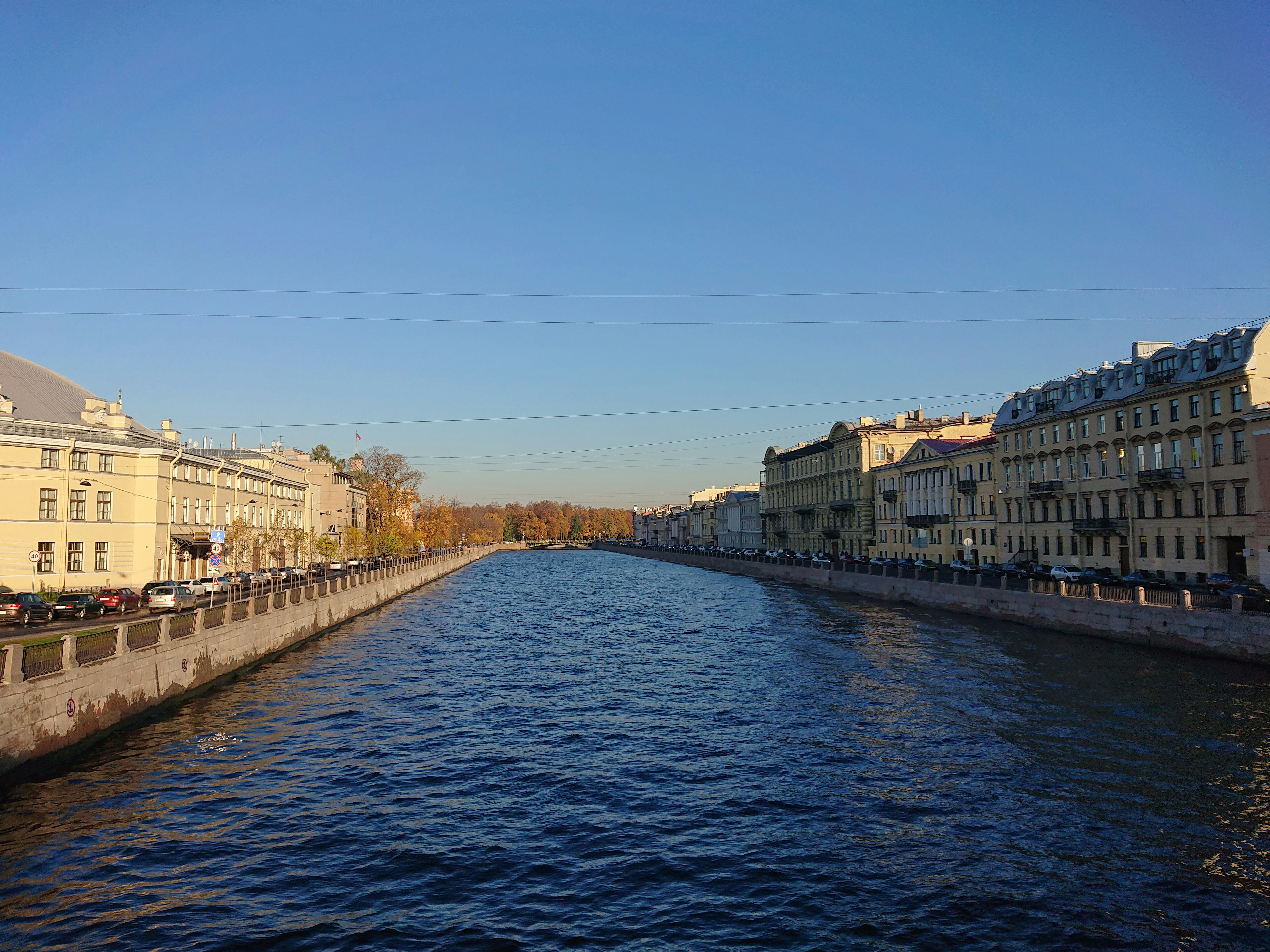 A serene view of a canal flanked by historic buildings under a clear blue sky, showcasing the tranquil waters and autumn foliage along the banks.