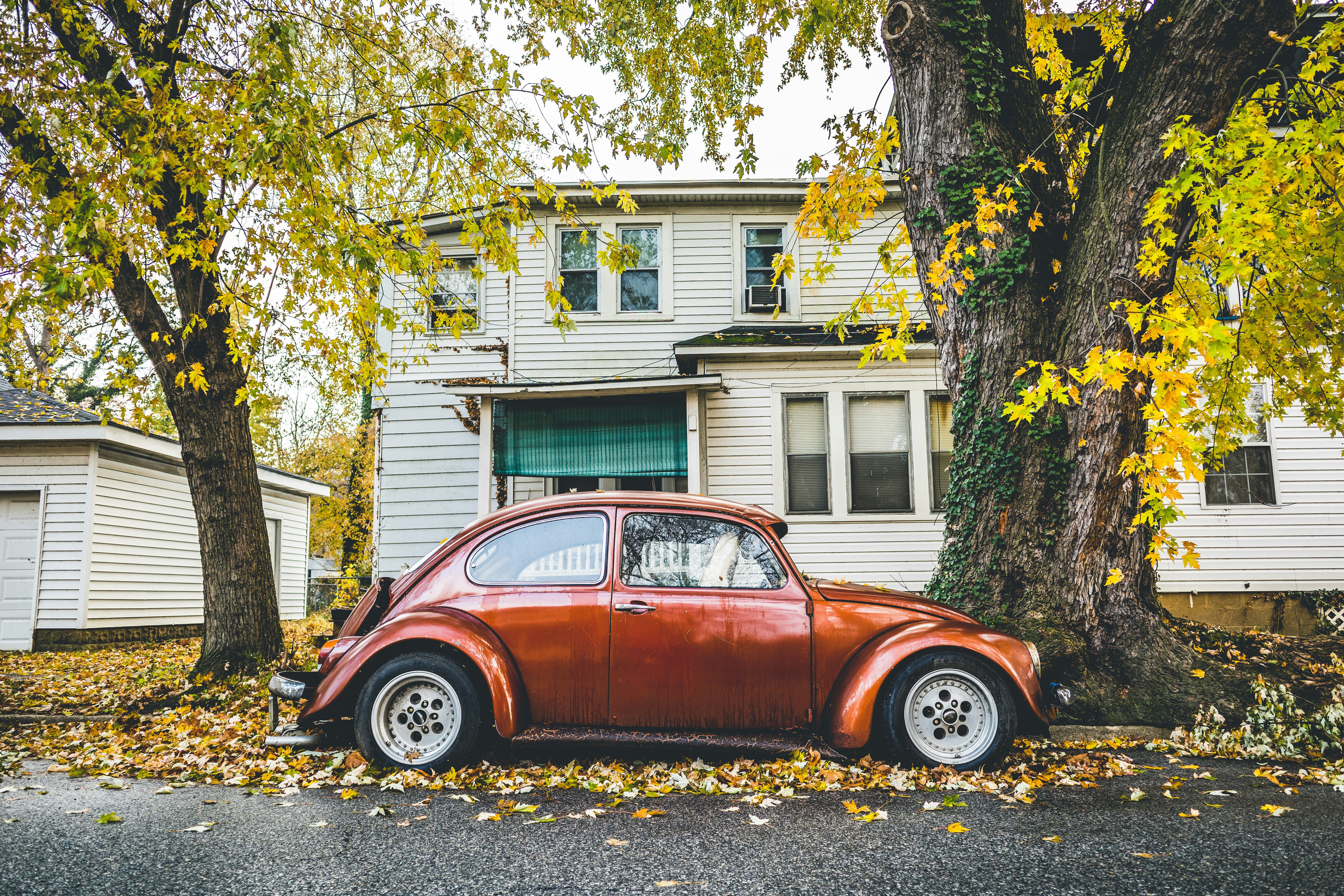 Classic red car parked under a tree with autumn foliage in front of a white house.