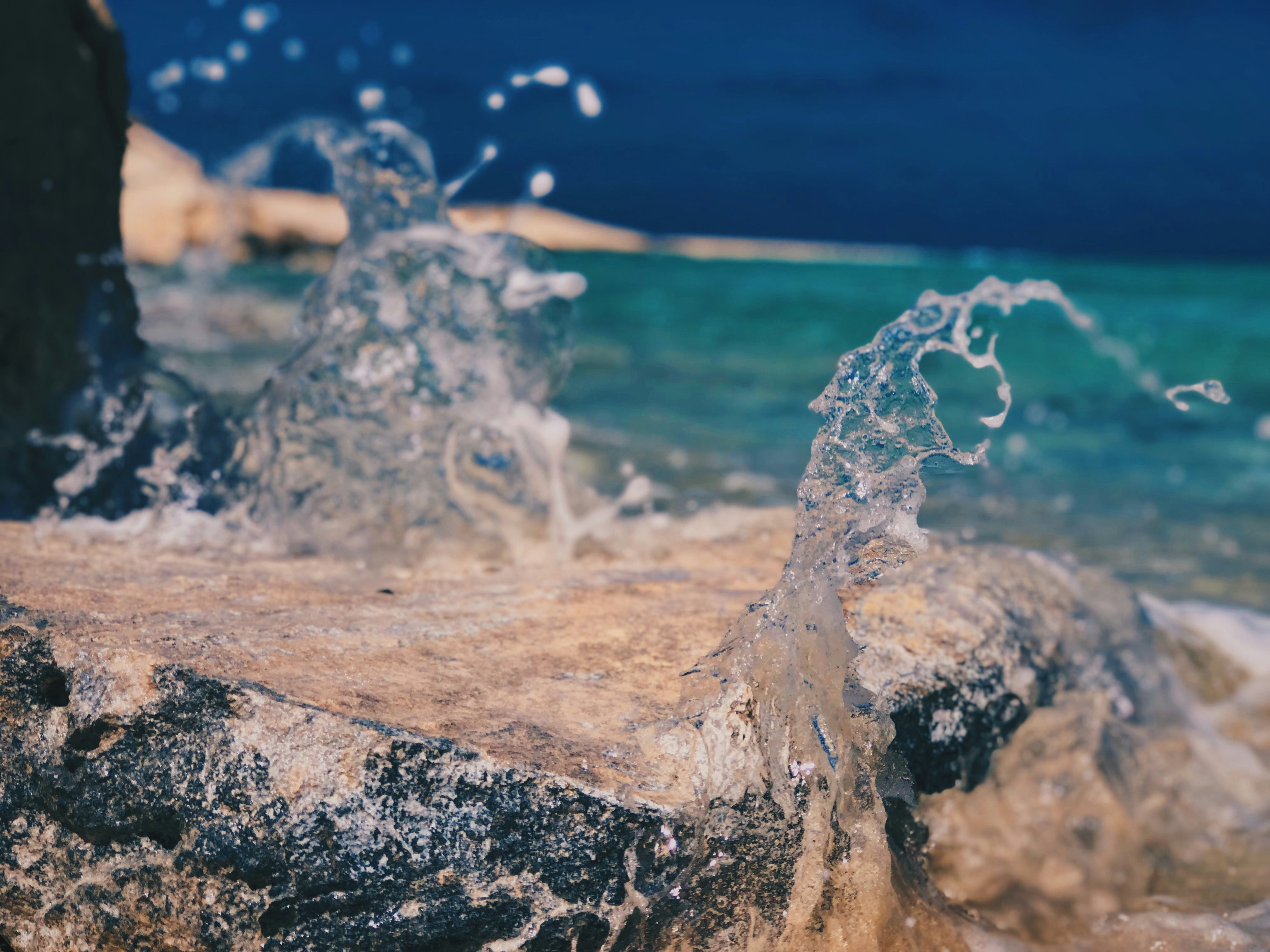 Waves crashing against rocks with water splashes frozen mid-air under a bright blue sky.