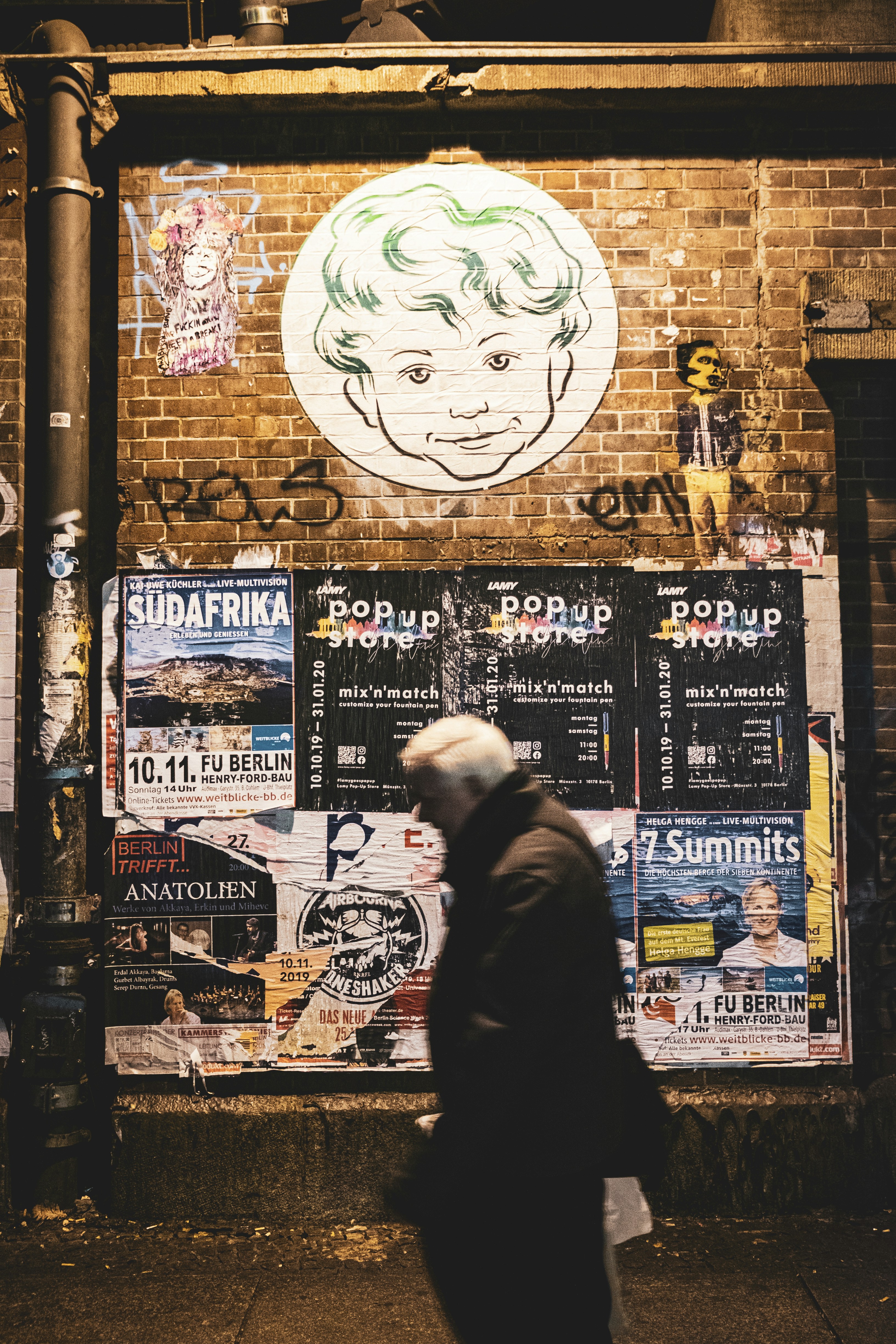 Nighttime mural scene featuring a large circular baby-face painting on a brick wall covered with posters, with a blurred passerby in the foreground.