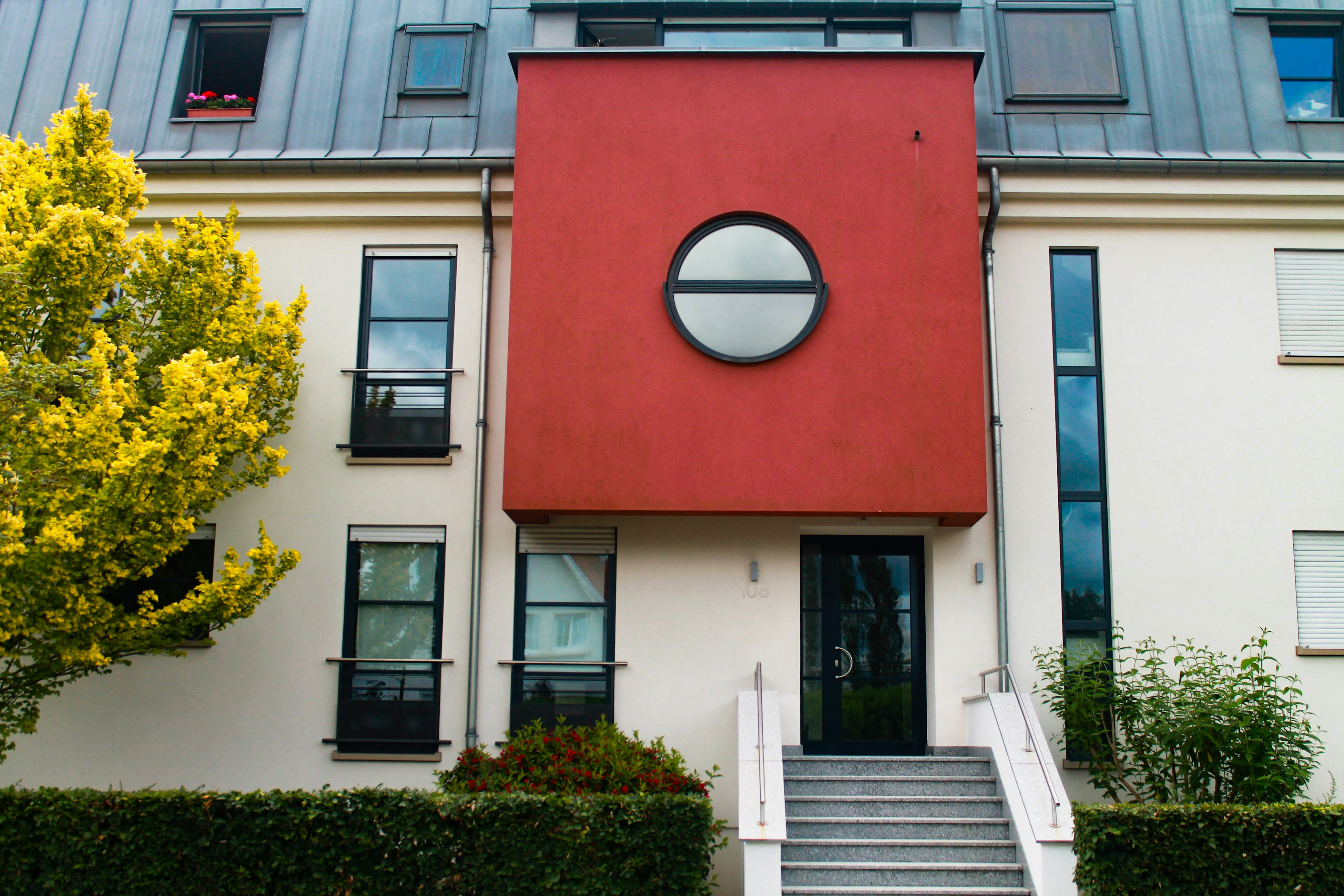 Contemporary building exterior featuring a bold red panel with a circular window, complemented by lush greenery and a staircase entrance.