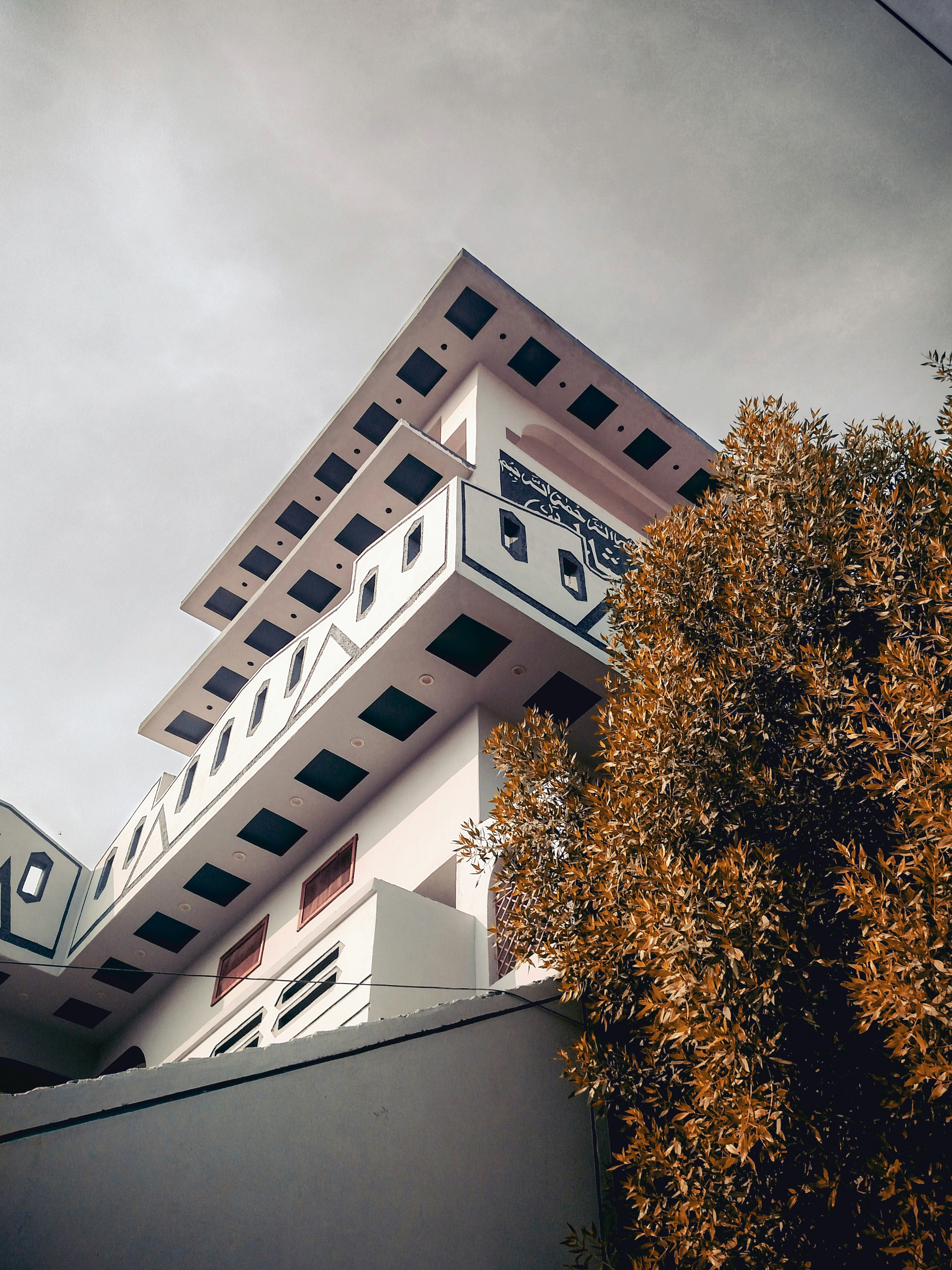 Architectural photograph of a stacked, overhanging building with bold geometric windows. A dense tree with autumn leaves frames the right side.