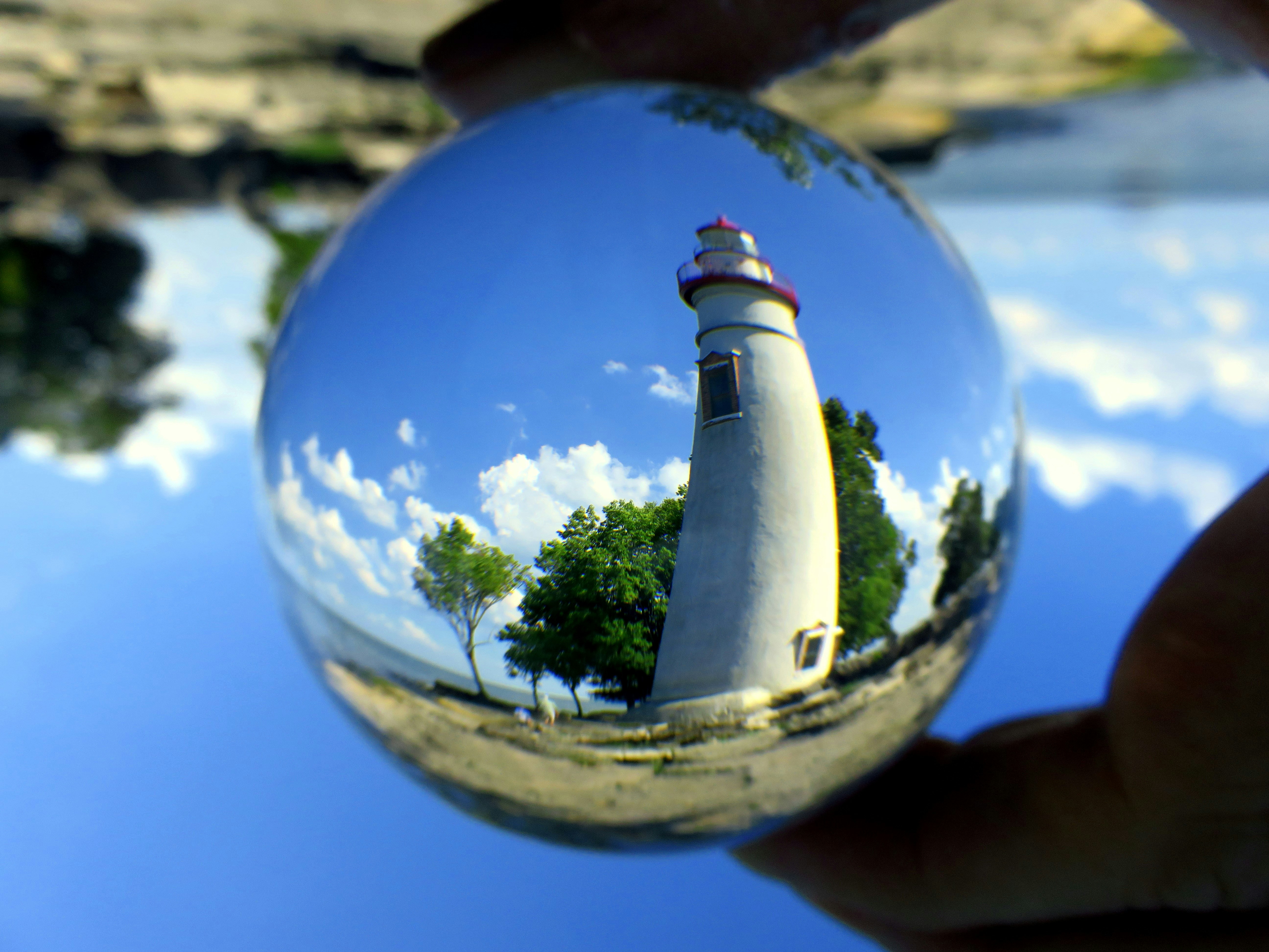White lighthouse viewed through a handheld glass sphere, inverting the surrounding landscape.