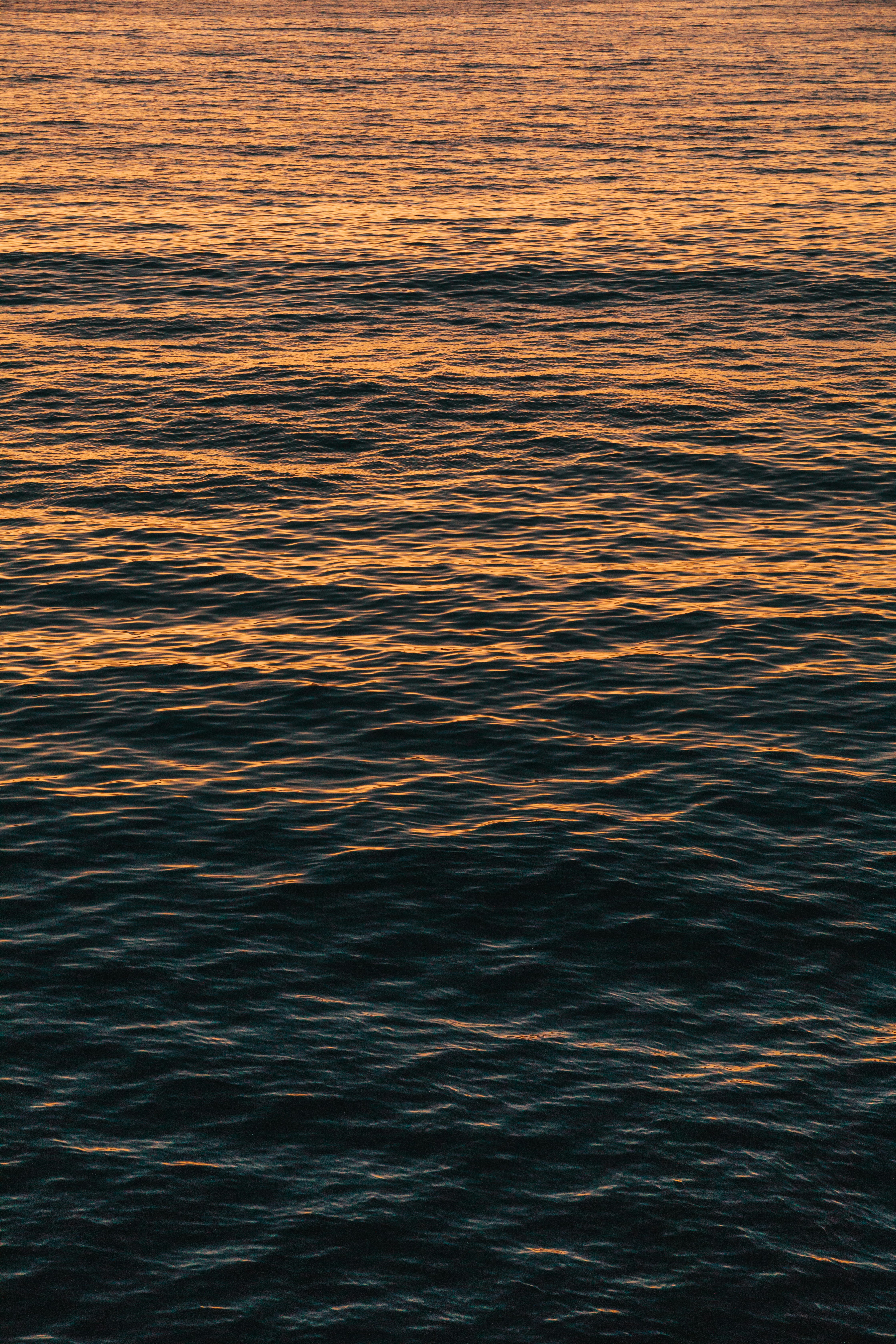 Gentle waves reflecting warm hues of sunset, creating a serene seascape. The surface glistens with soft, rippling patterns.