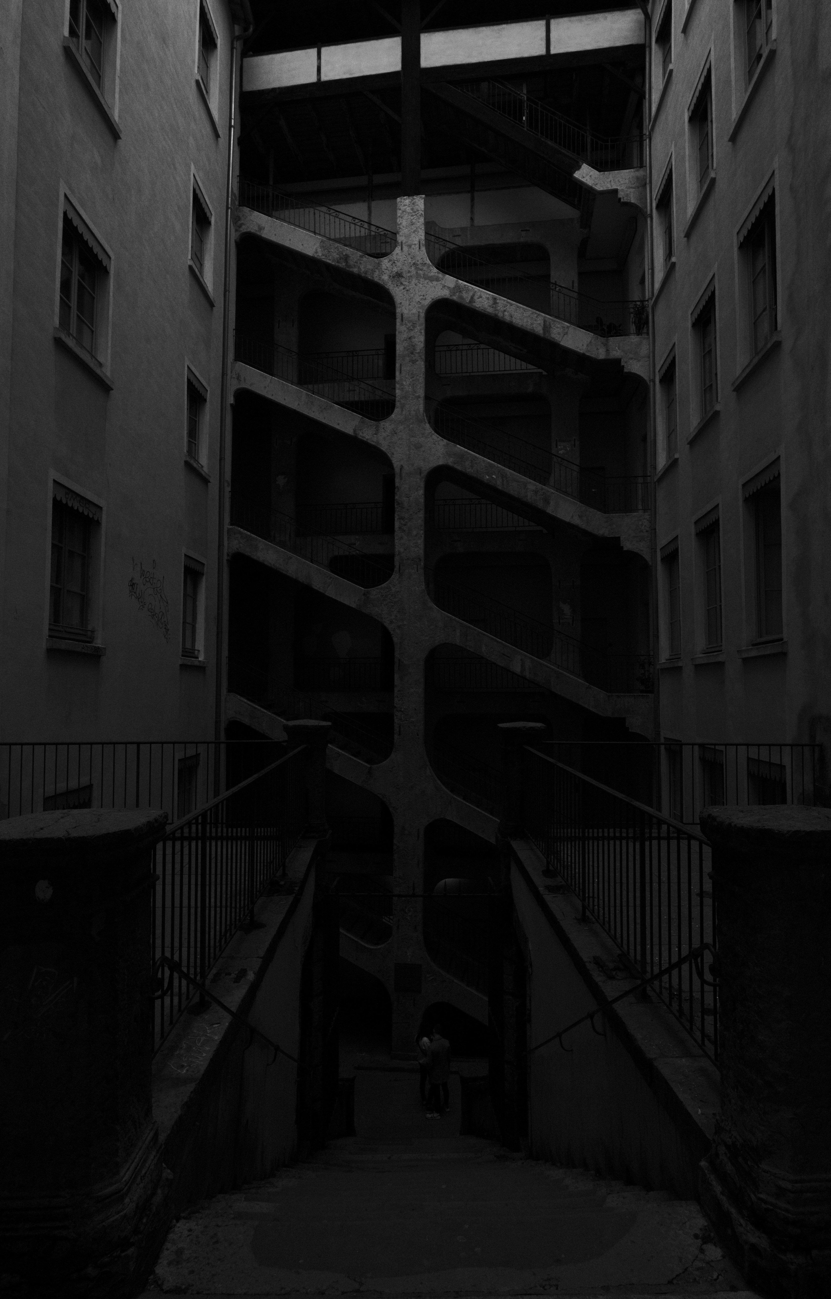 A stark, monochromatic view of a winding staircase framed by two buildings, evoking a sense of isolation. A lone figure stands at the base, emphasizing the scale and architectural lines.