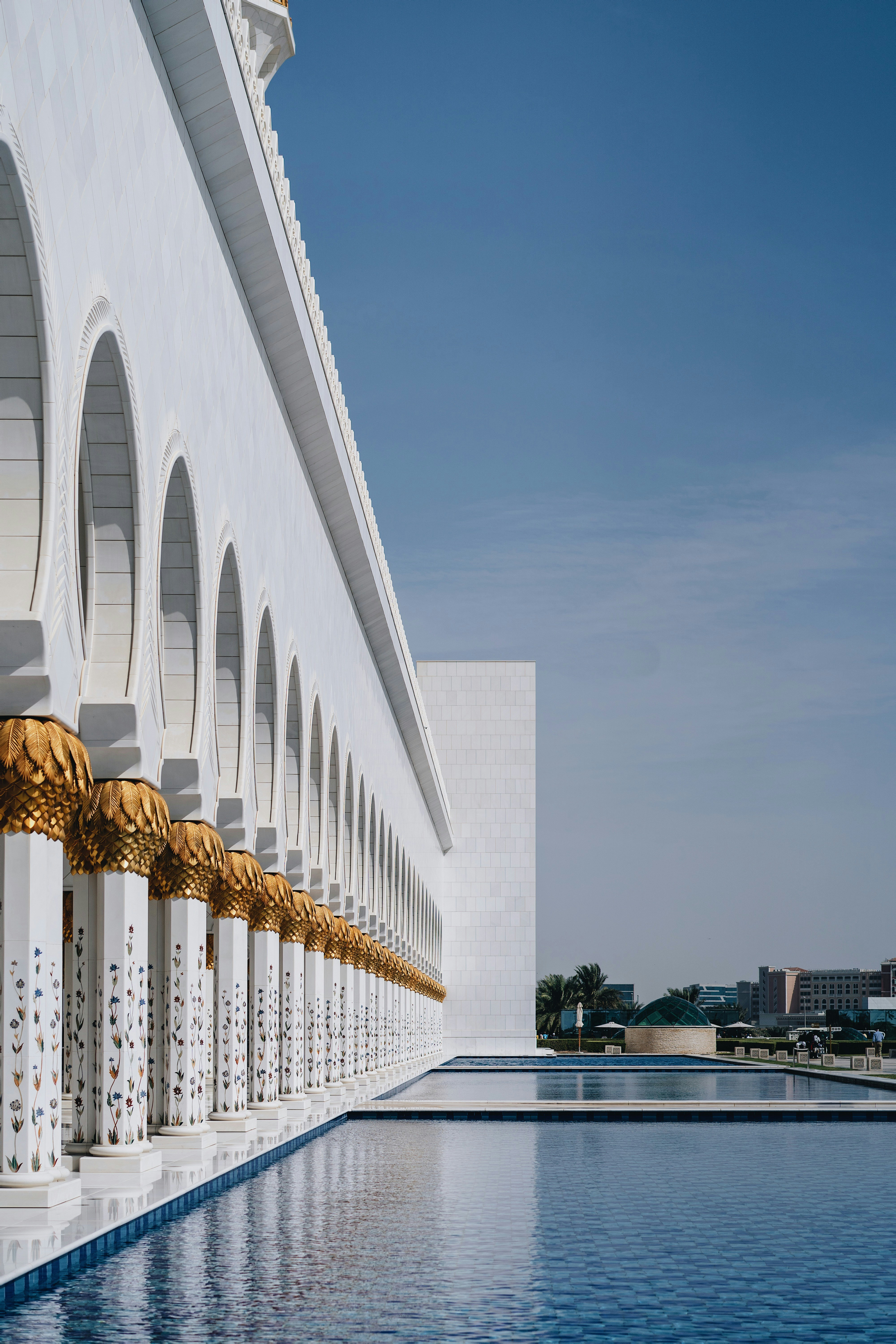 Elegant white marble arches adorned with golden accents, reflecting in a tranquil pool under a clear blue sky.